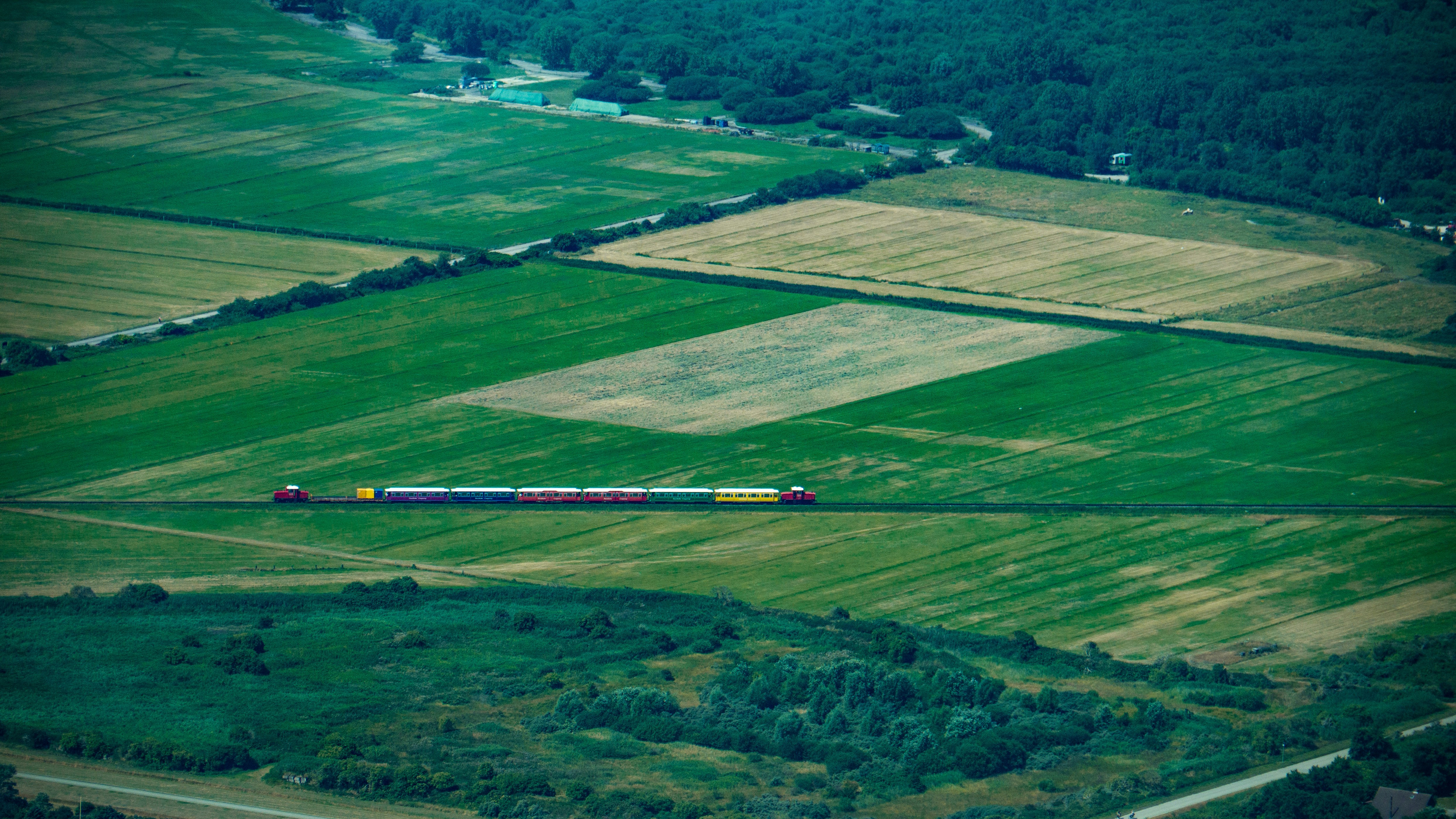 Freight train traversing vibrant green fields and agricultural patches, showcasing the contrast of colors and textures in the landscape.