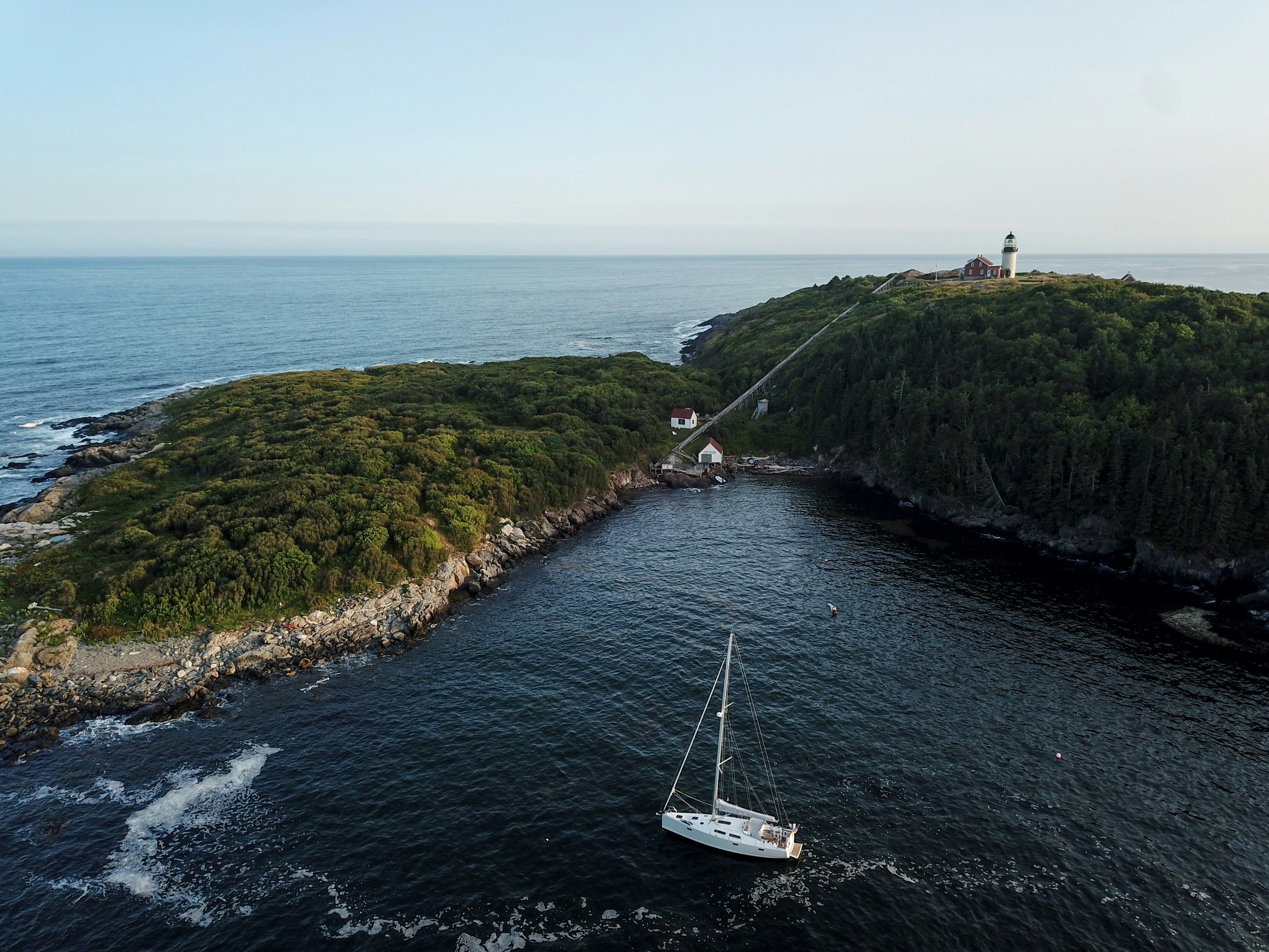 Sailboat in a Quiet Cove Framed by Forested Headlands and a Distant Lighthouse