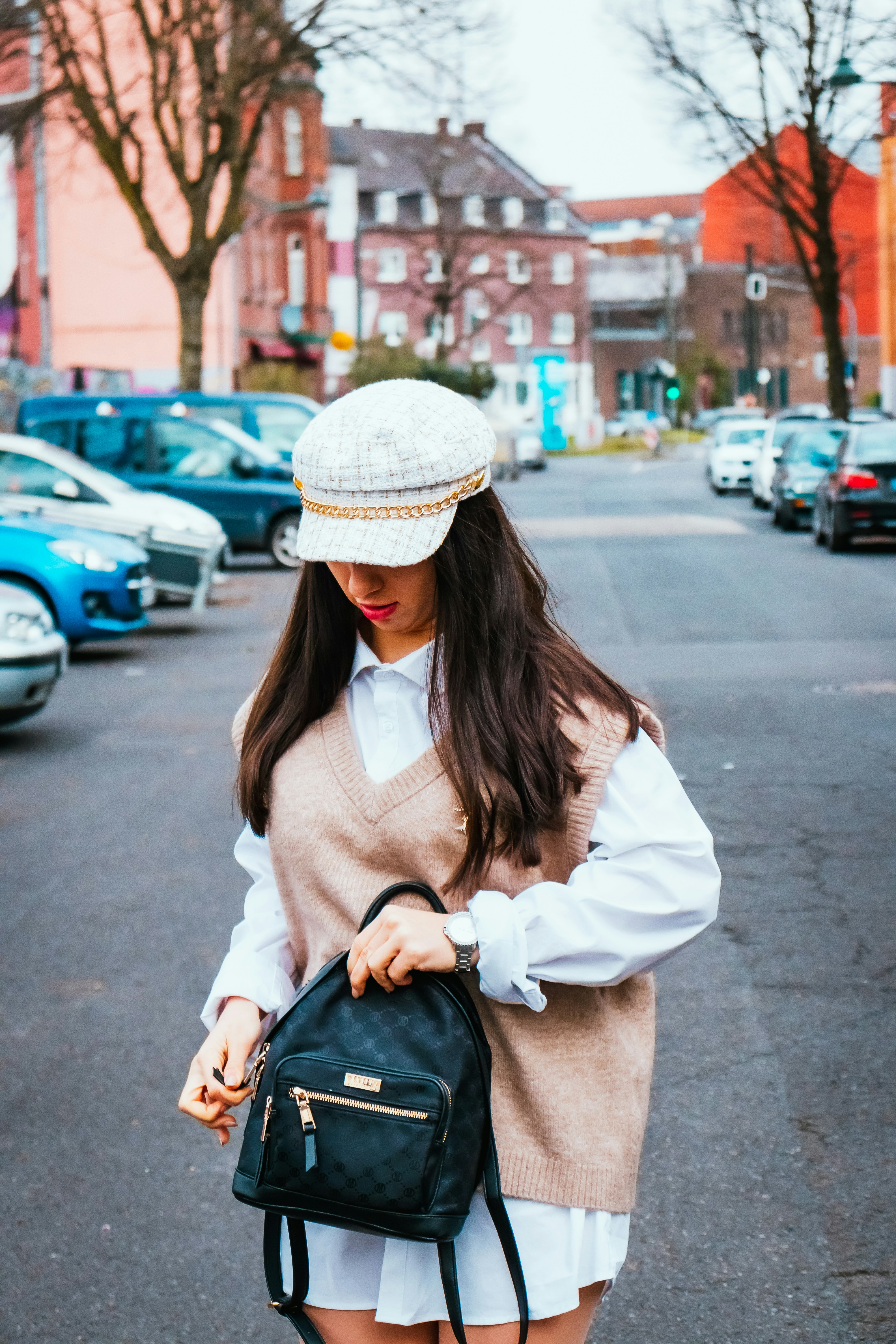 A young woman in a stylish outfit featuring a beige vest and a cap, holding a black backpack while walking through an urban street.