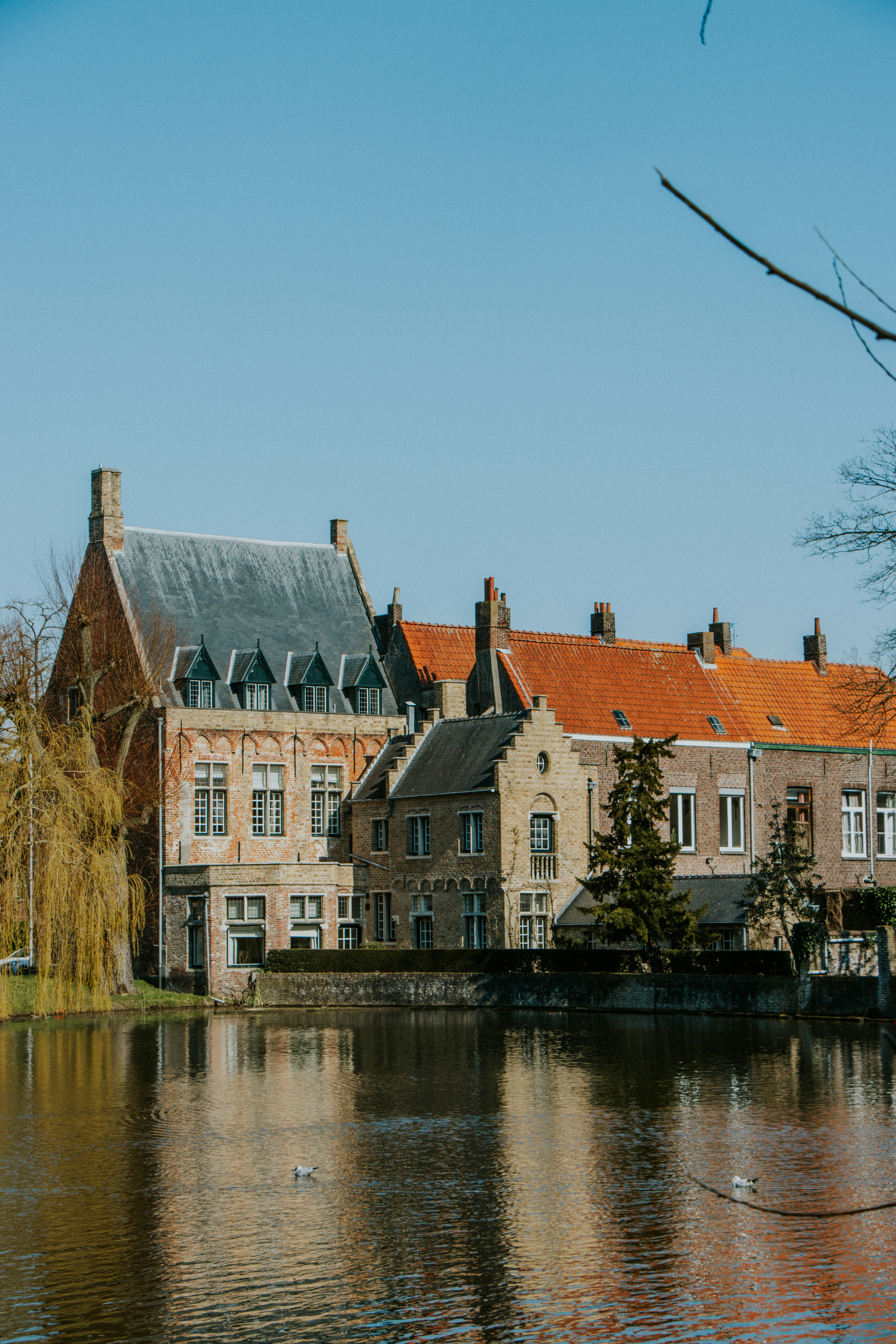 Historic brick buildings with distinctive roofs reflecting in calm waters, framed by trees and a clear blue sky.