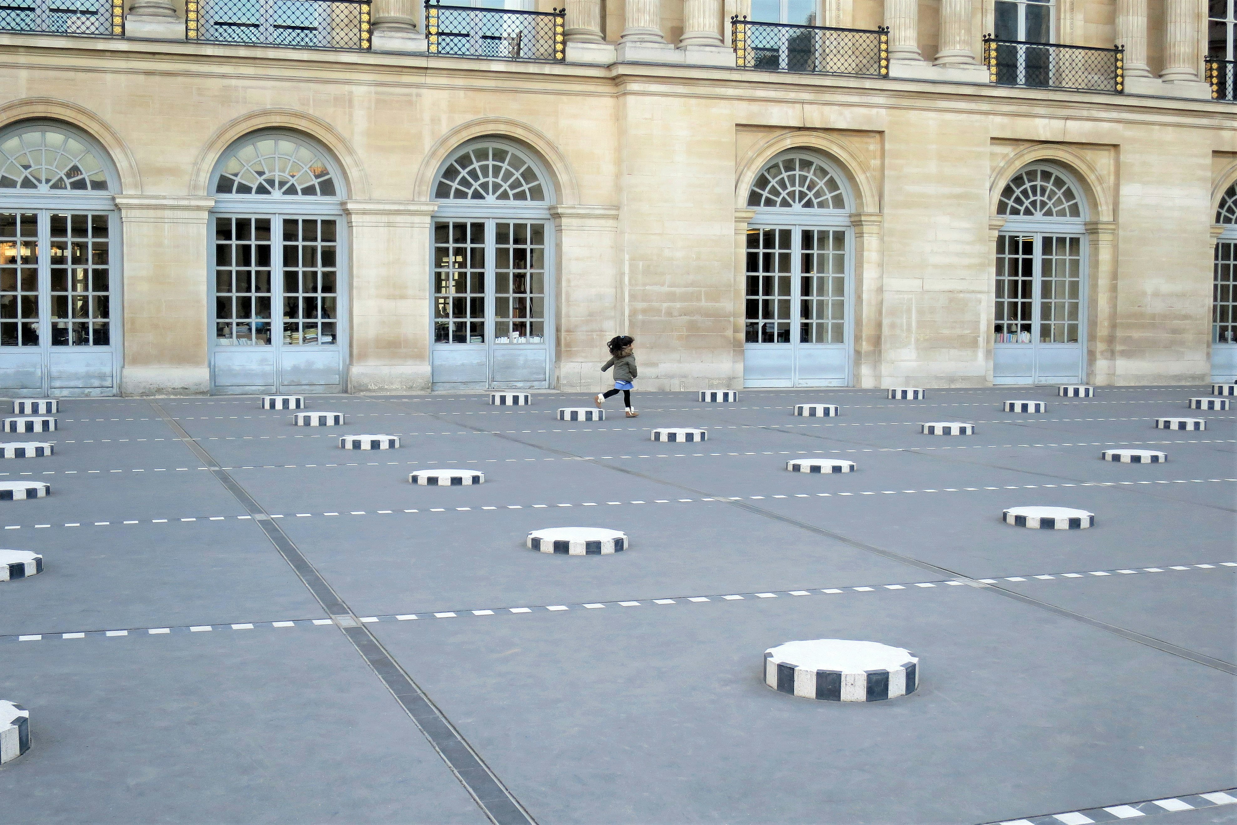 A child joyfully runs across a patterned plaza, surrounded by circular installations and elegant architecture. The scene captures a blend of movement and design.