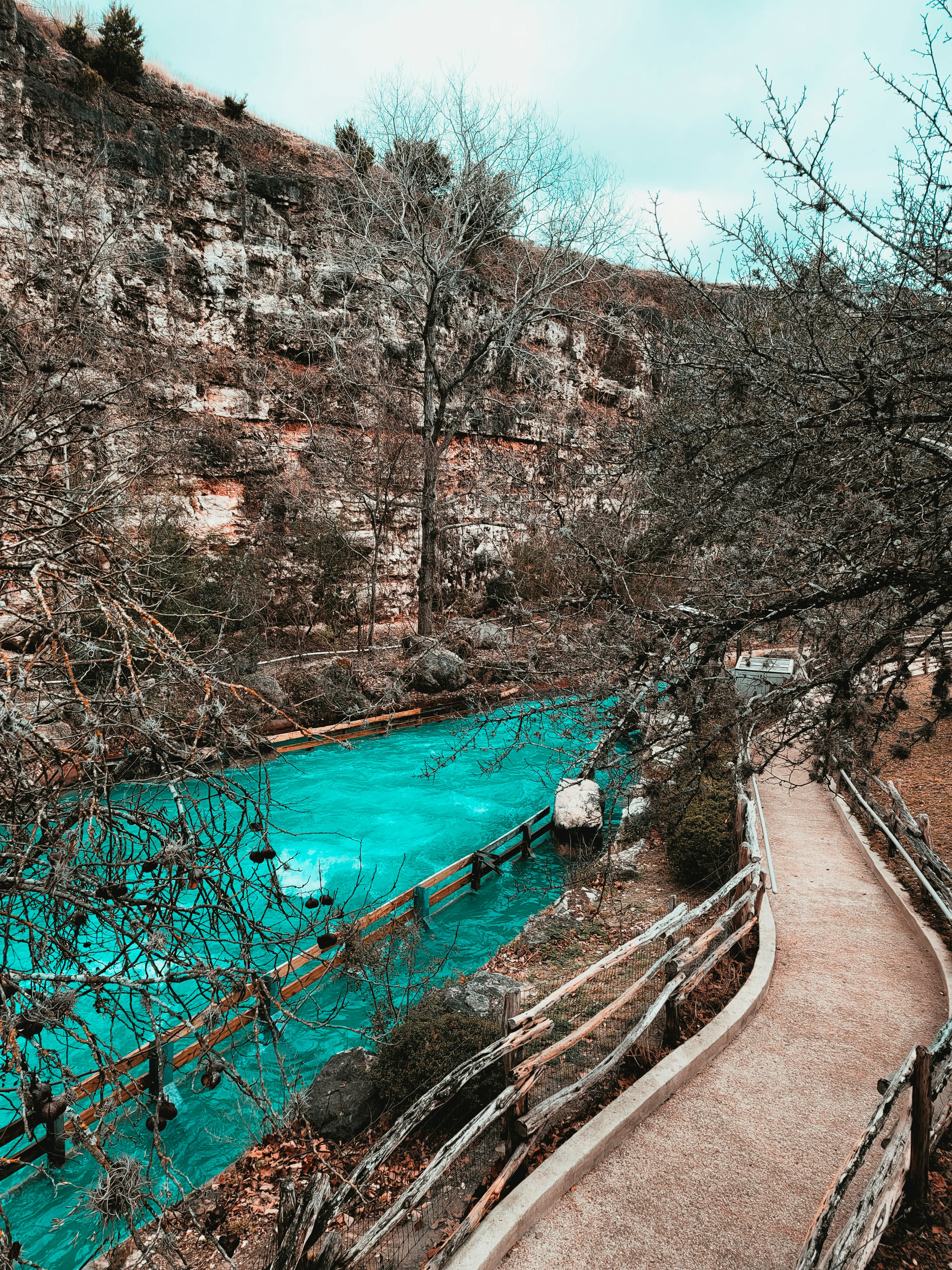 Colorful turquoise pool beside a winding boardwalk carved into a rocky cliff, with bare trees and rugged terrain in the background.