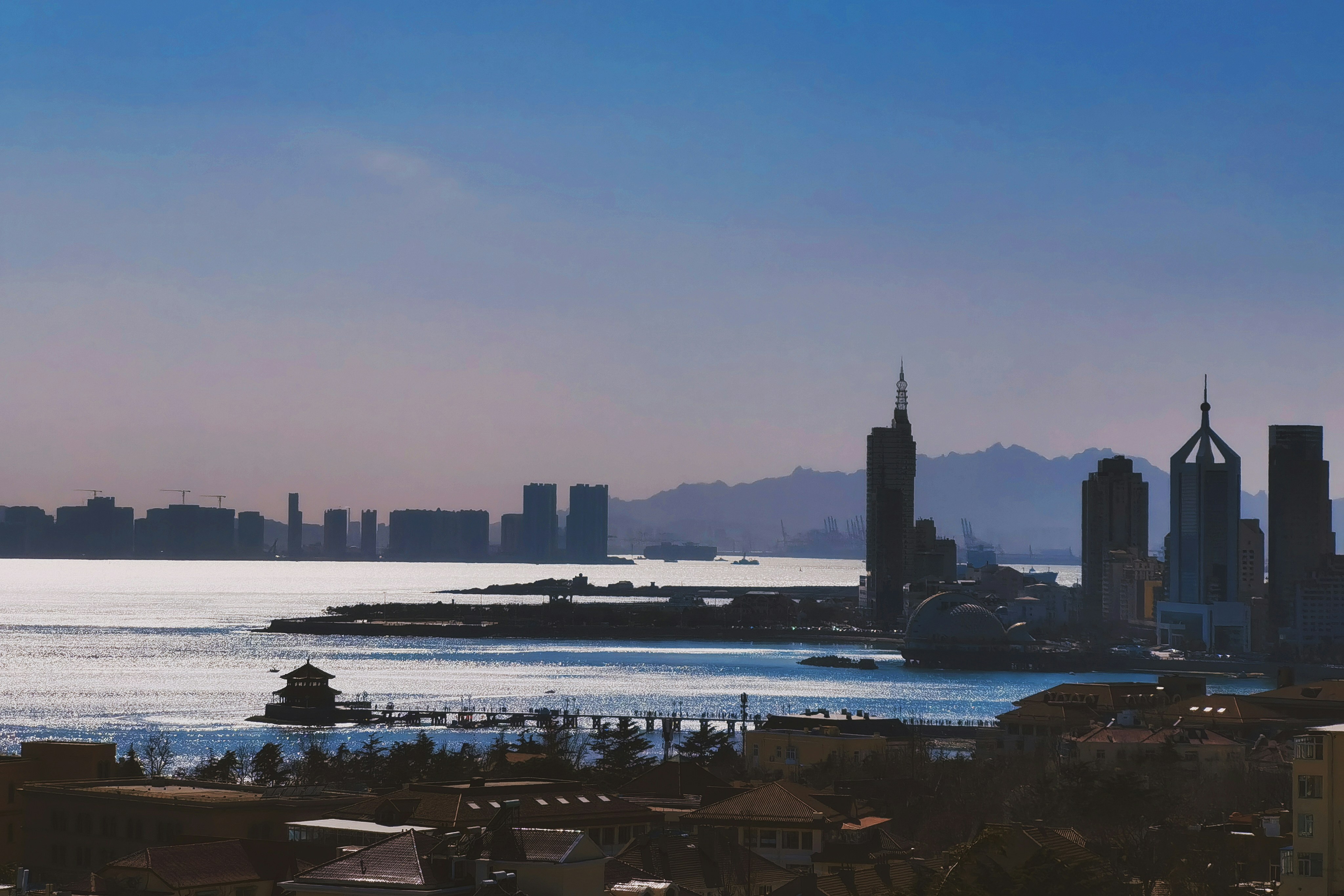 City skyline silhouetted against a twilight sky with distant mountains and shimmering waters.