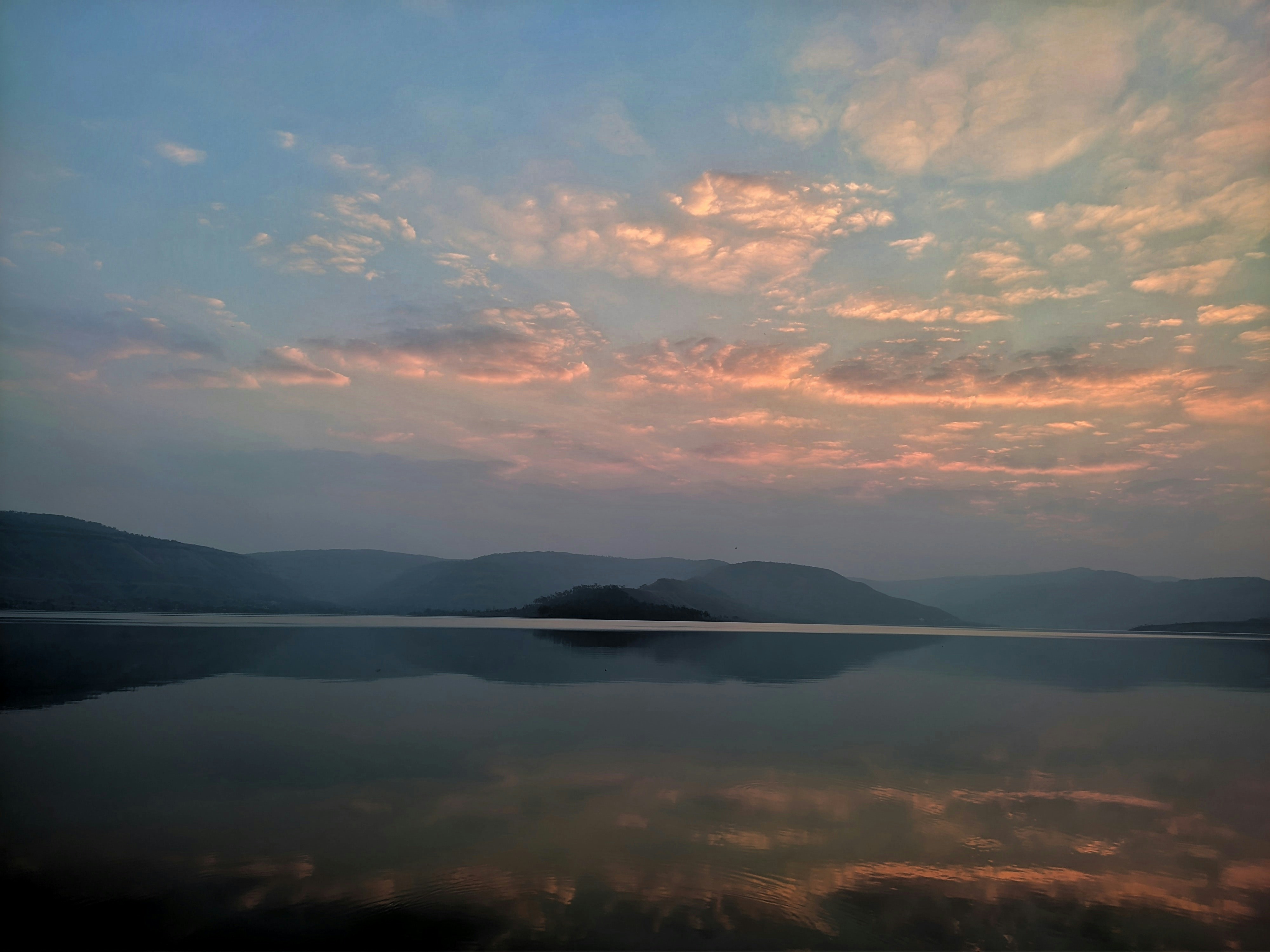 Soft pastel clouds mirror in a tranquil lake surrounded by distant hills at dusk.