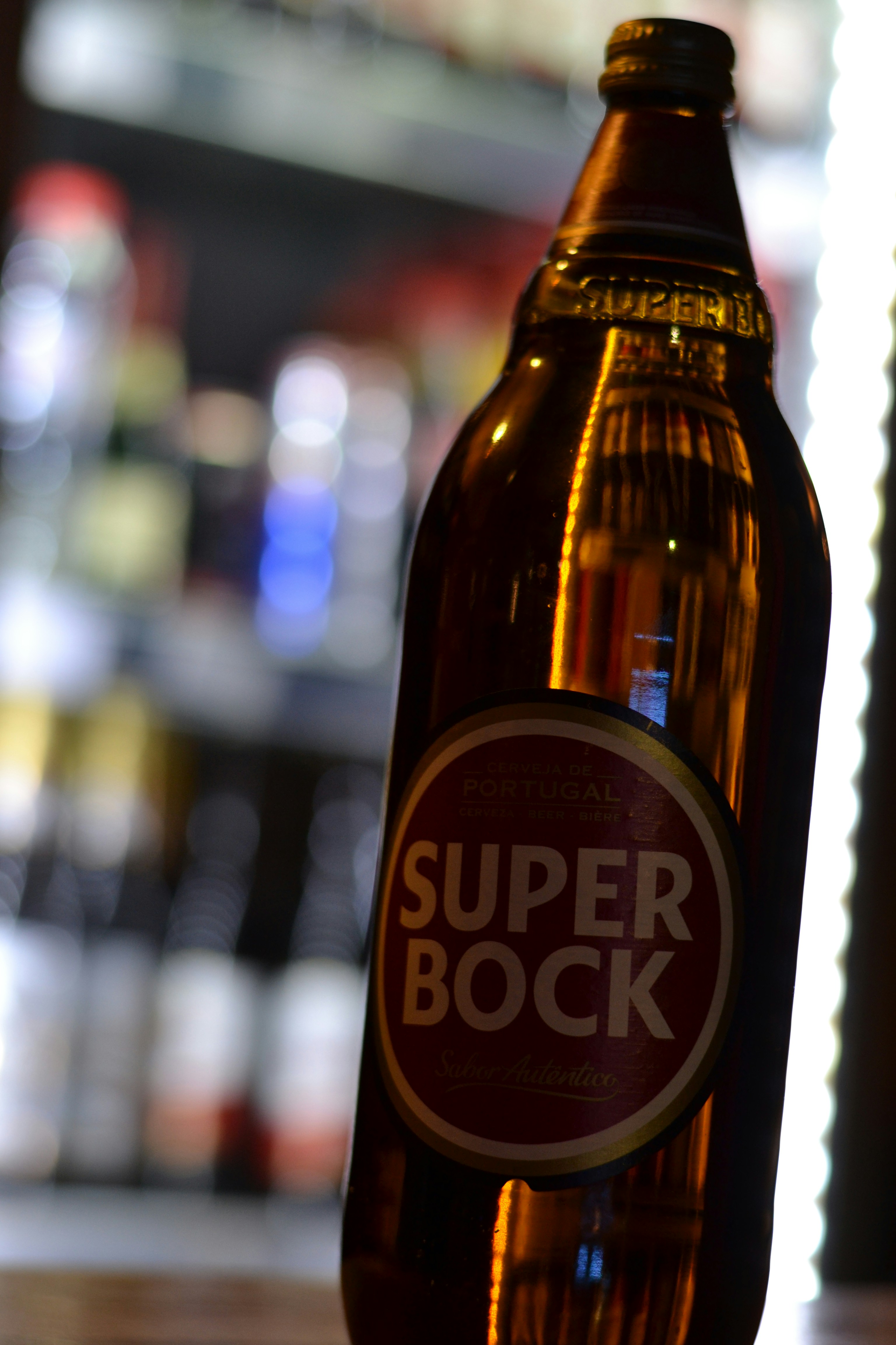 A close-up of a Super Bock beer bottle, illuminated against a blurred backdrop of bar shelves filled with various drinks.