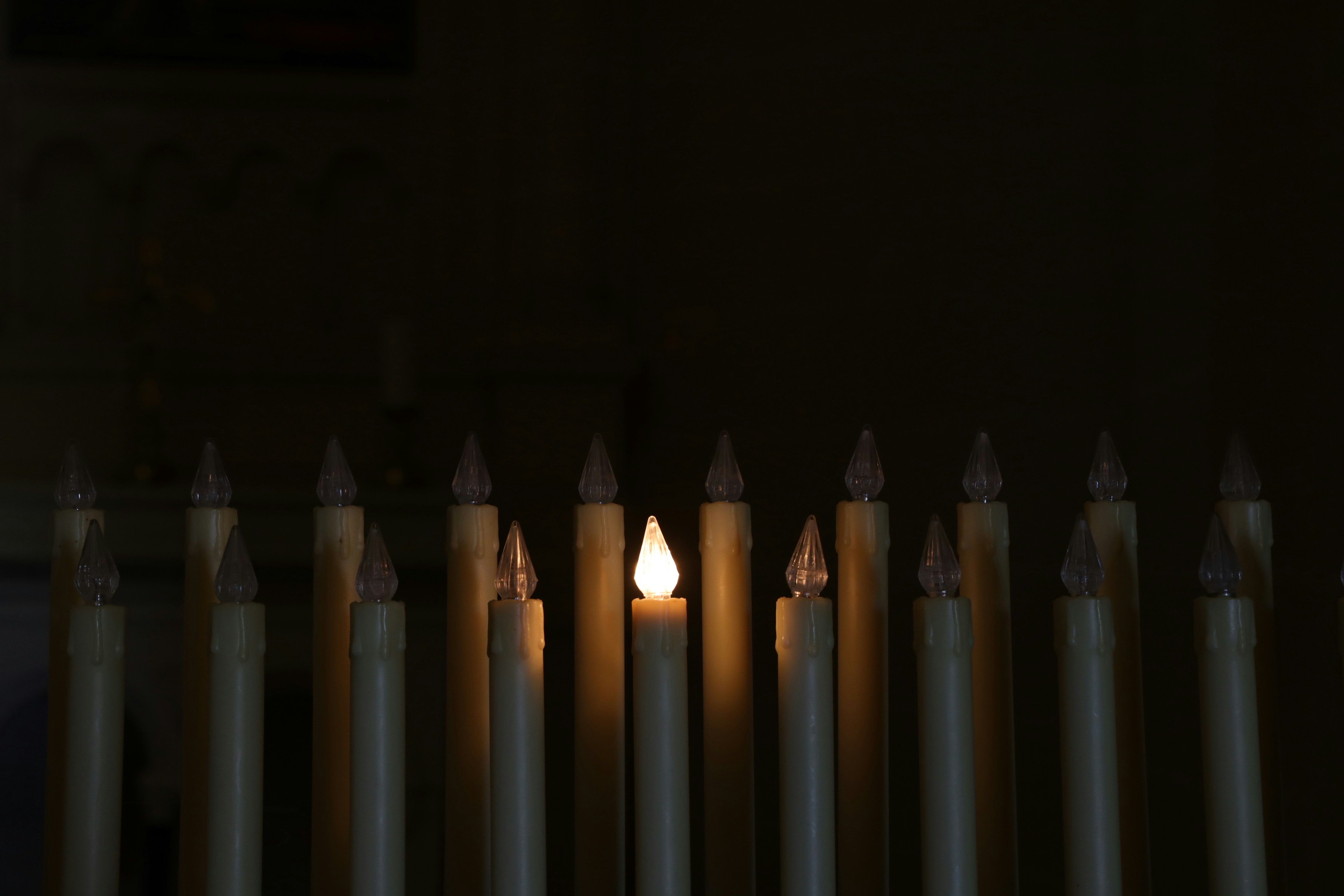 A single lit candle stands out among a row of unlit candles, creating a striking contrast in a dimly lit setting.