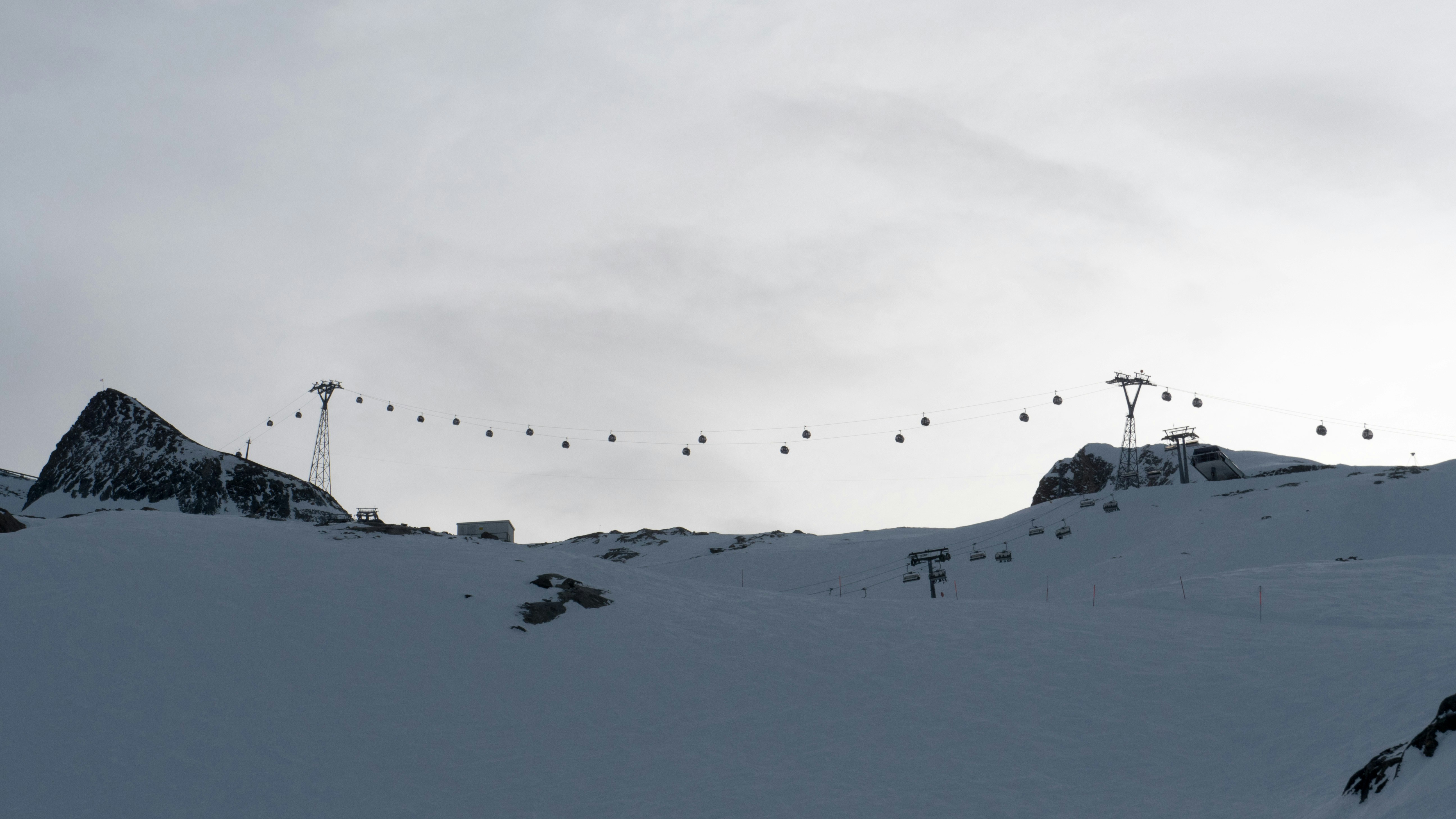 Ski gondolas traverse a snowy landscape, silhouetted against a soft, overcast sky. The stark contrast emphasizes the serene, wintry environment.