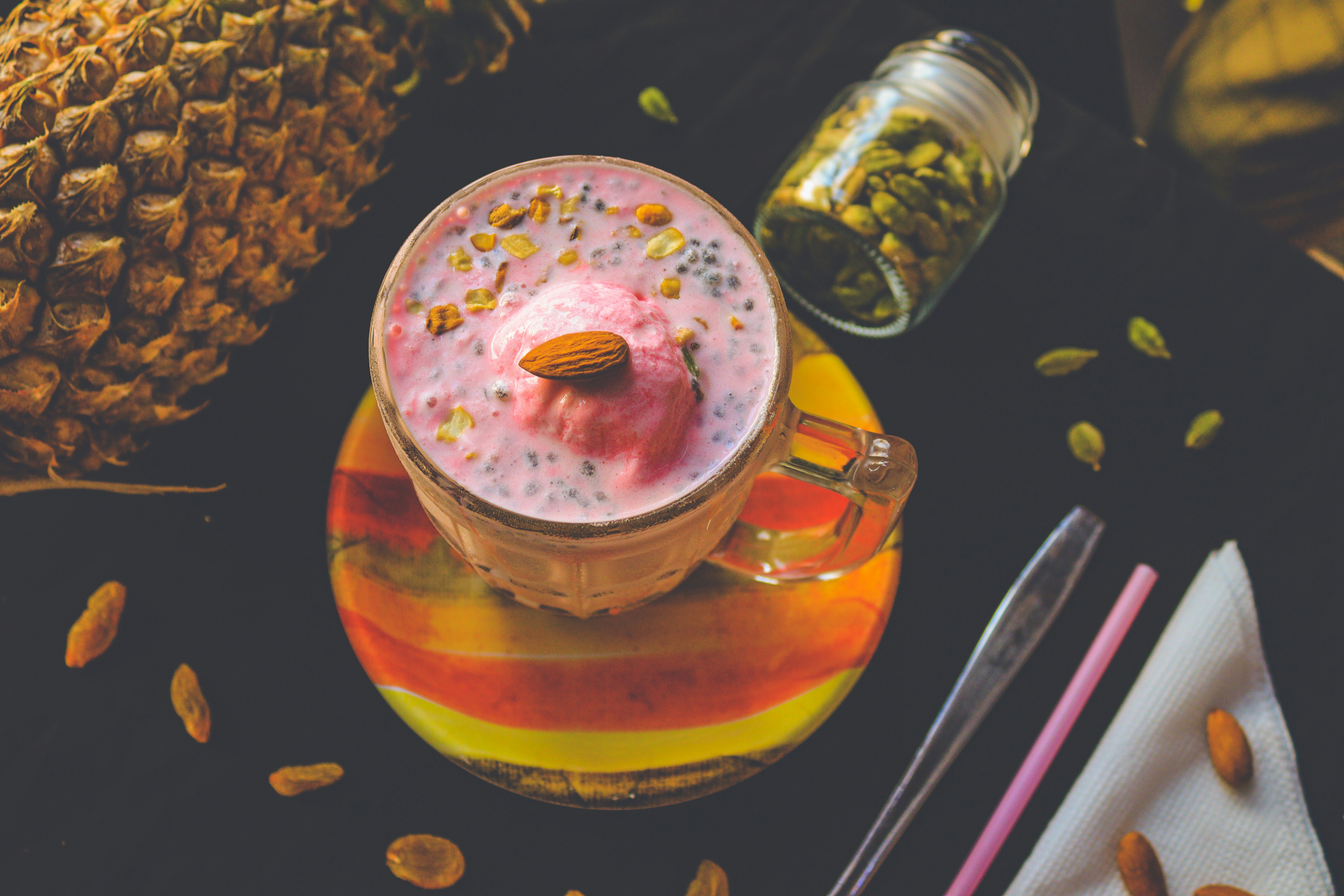Colorful beverage topped with nuts alongside scattered spices and a pineapple on a dark surface.