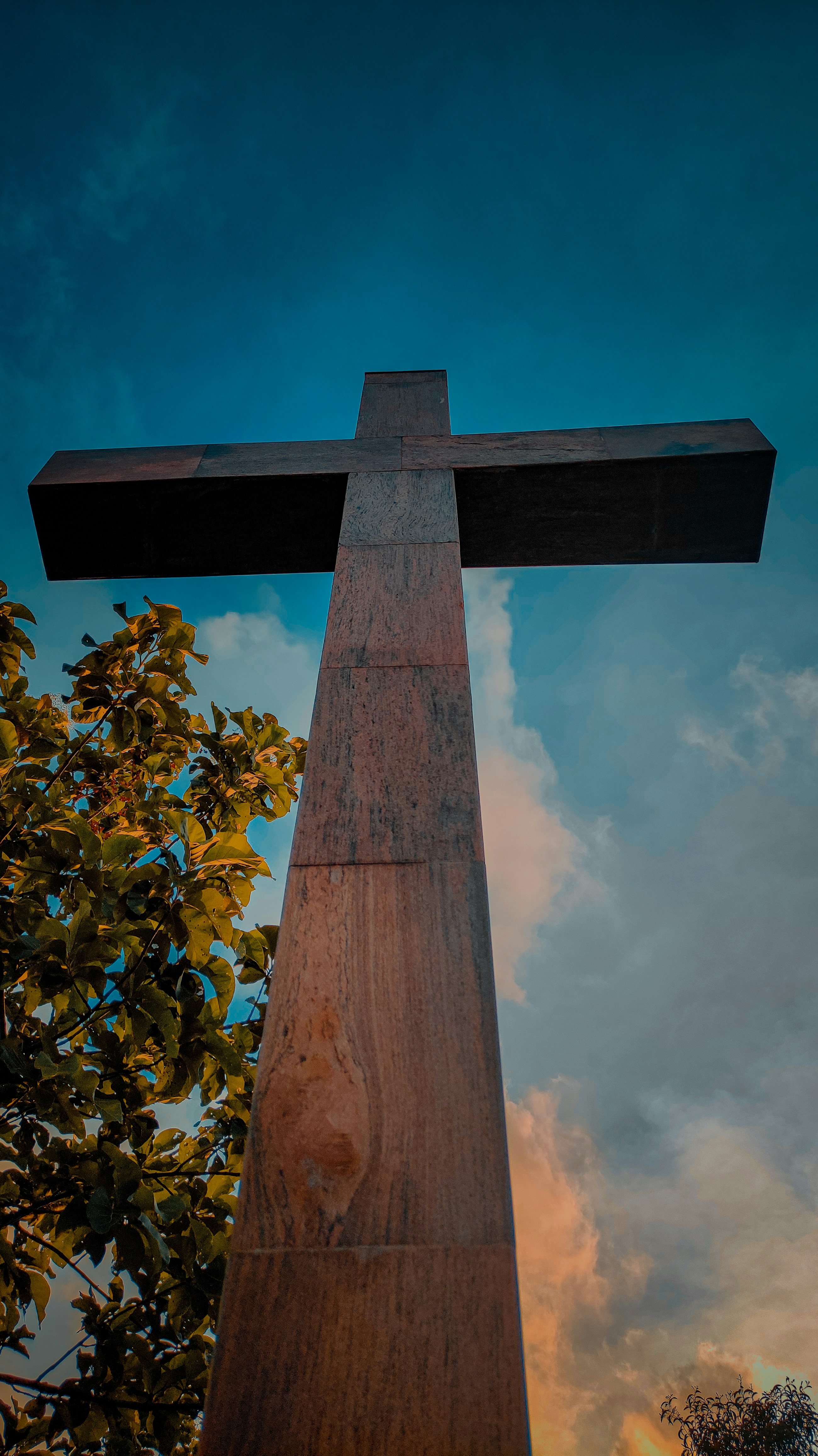 A towering wooden cross framed by lush green leaves under a dramatic sky at dusk.