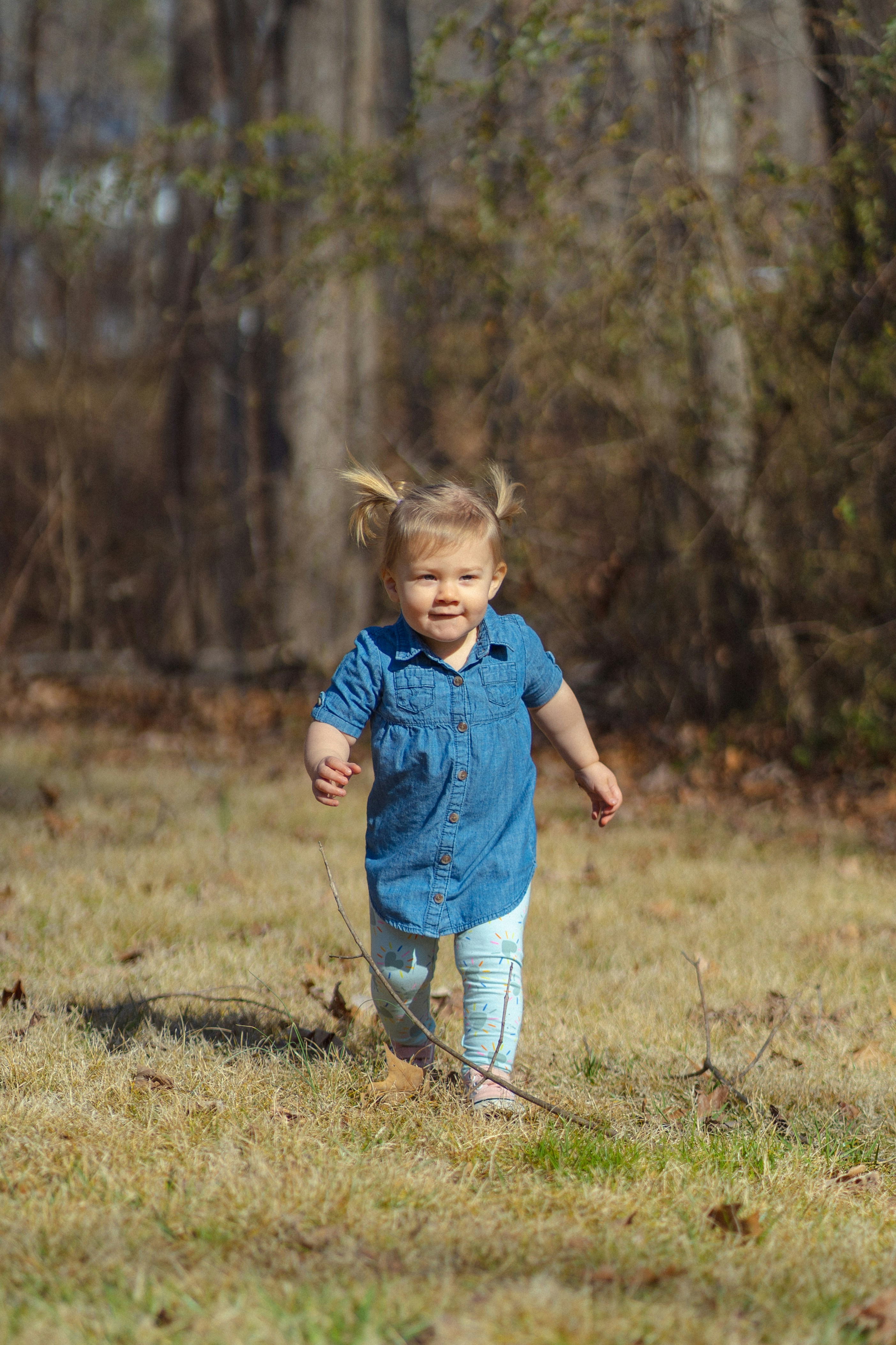 Toddler in denim shirt joyfully running through a grassy field, surrounded by trees in the background.
