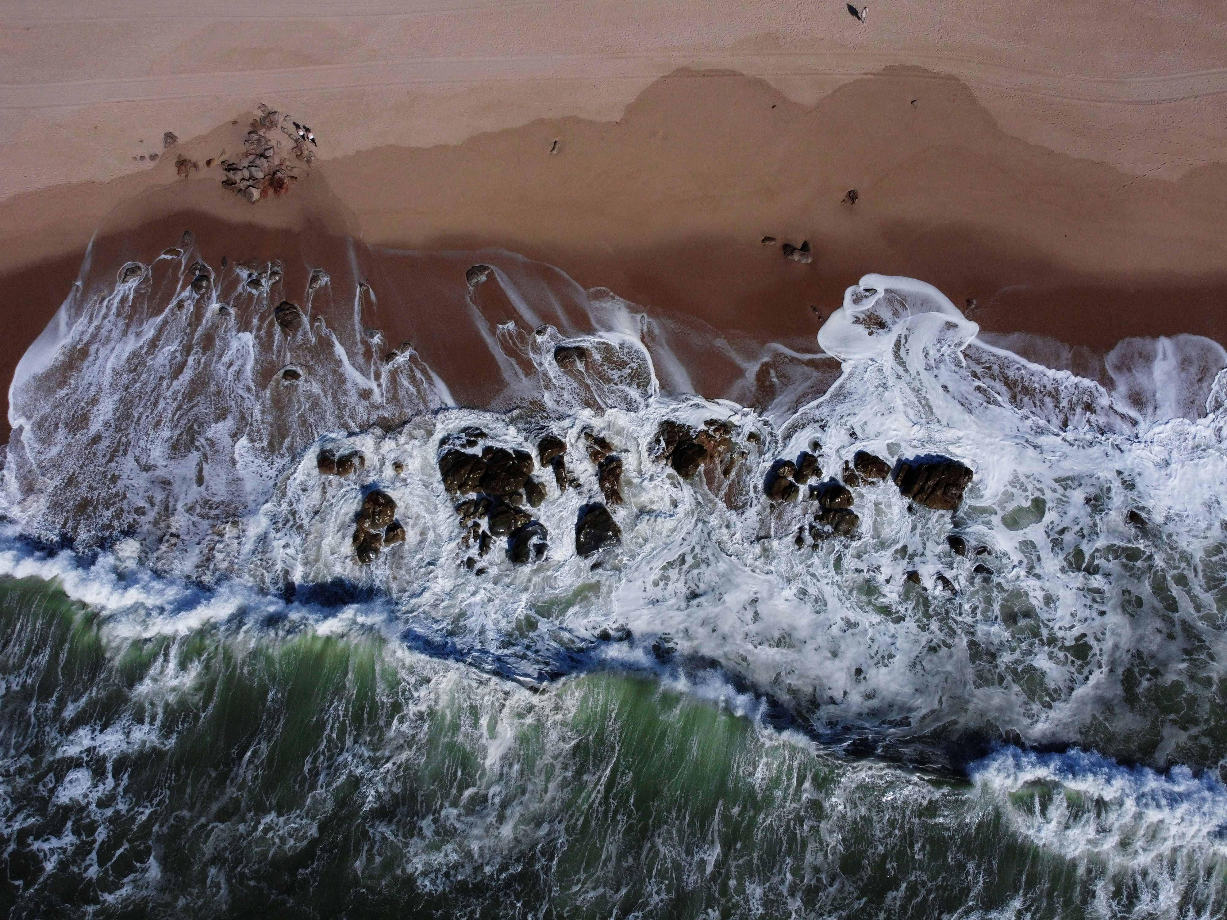 Foamy waves crashing against rocky outcrops on a sandy beach, captured from above. The interplay of water and land creates a dynamic coastal scene.