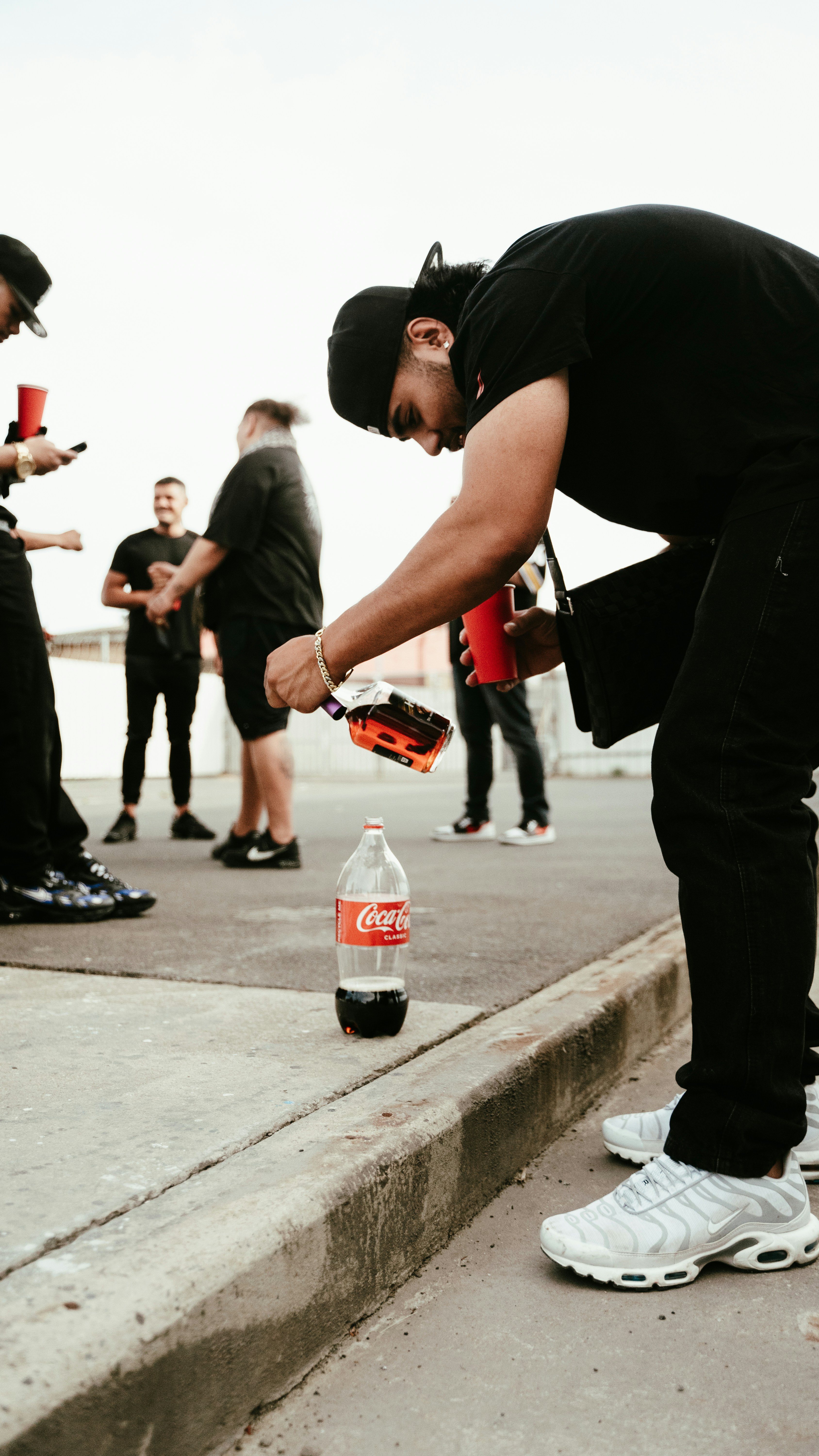 A group of friends gathers around as one person pours a colorful liquid into a Coca-Cola bottle, showcasing a playful moment of mixing drinks.
