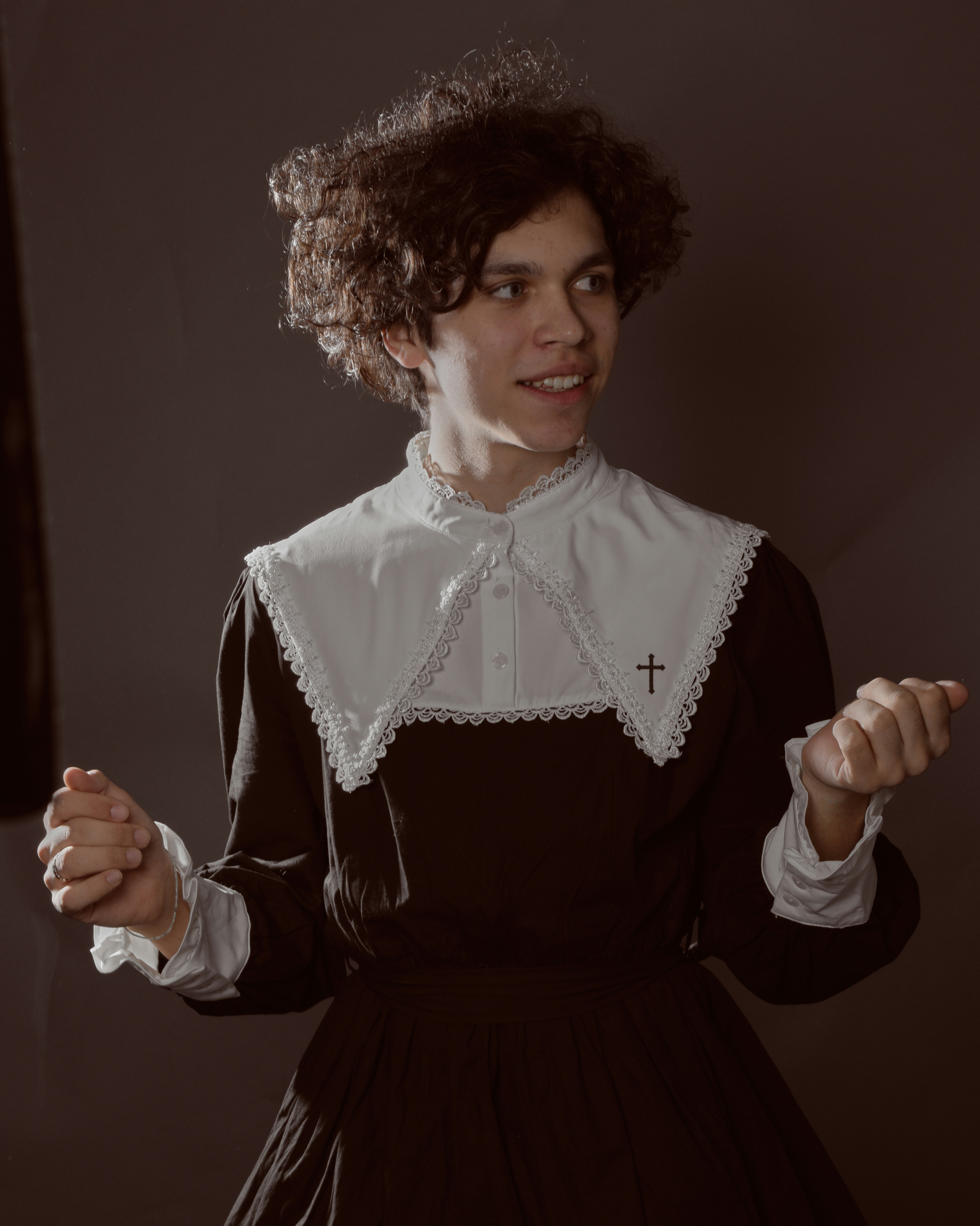 A young individual in vintage attire, featuring a lace-collared dress, poses with an expressive demeanor against a muted backdrop.