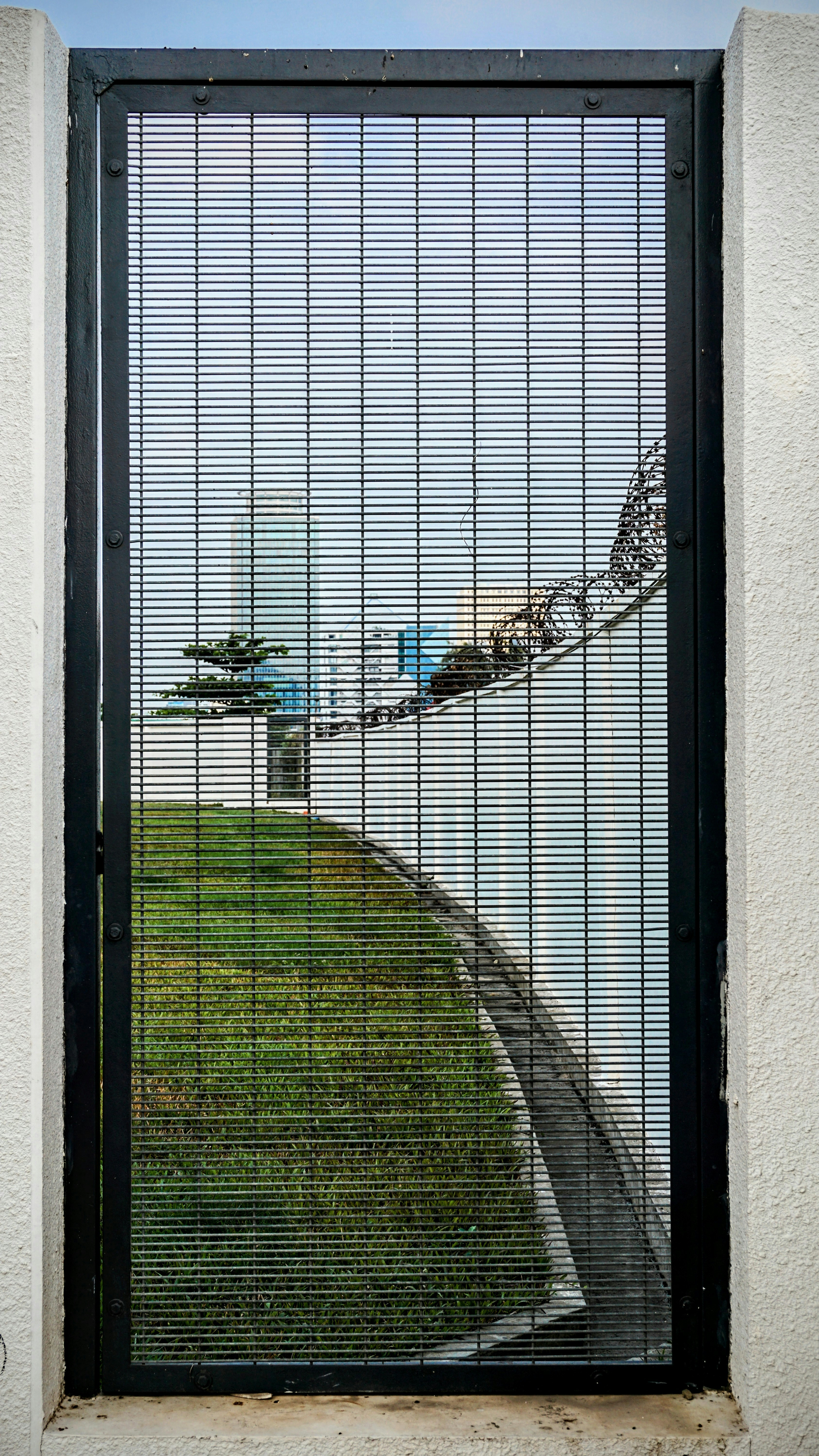 A view through a grated gate, revealing a curved pathway and urban landscape beyond. The foreground features a textured fence contrasting with the greenery and buildings in the background.