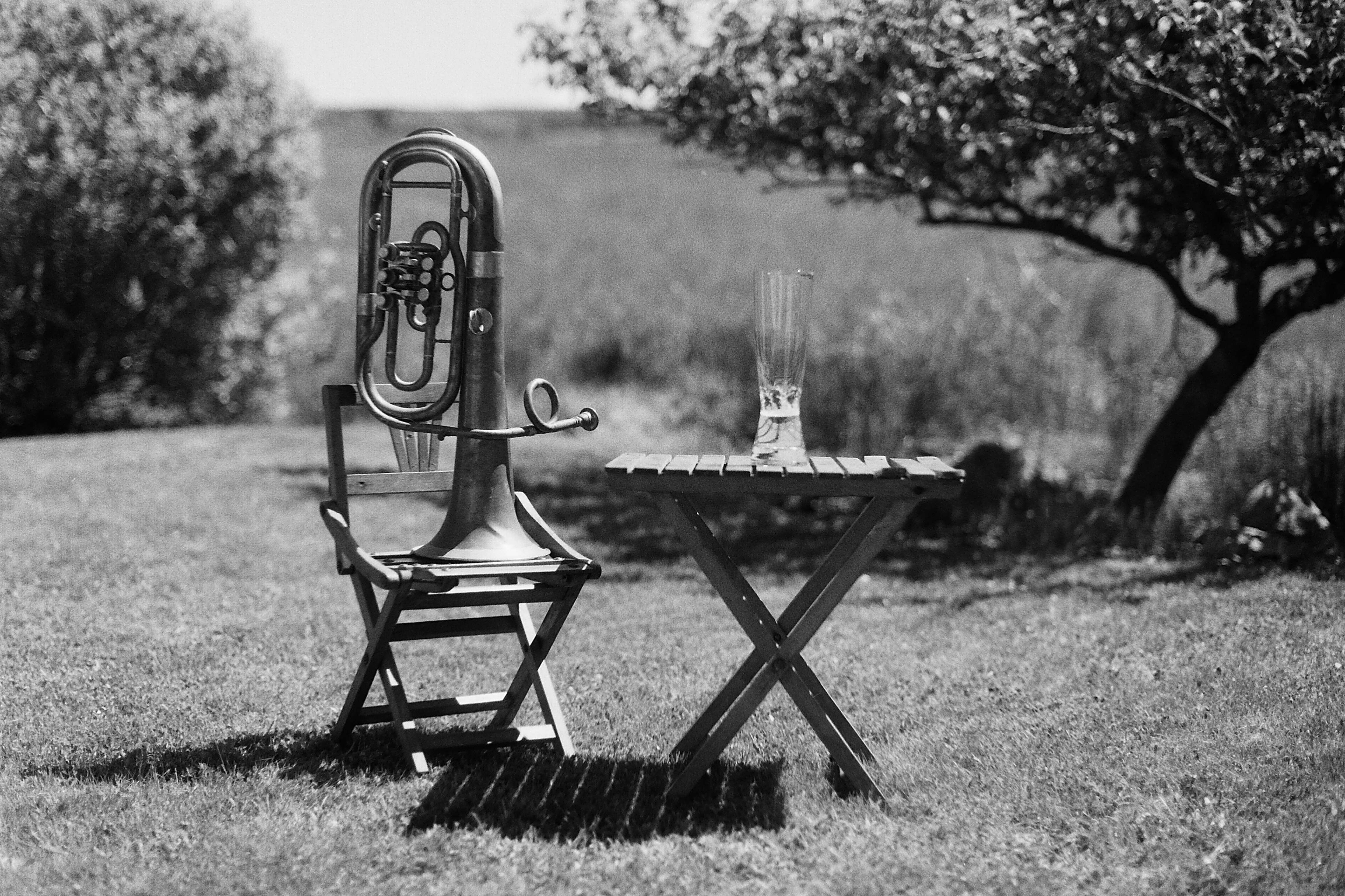 A tuba resting on a chair beside a small table with two glasses, set against a serene outdoor backdrop. The composition evokes a sense of leisure and musicality.