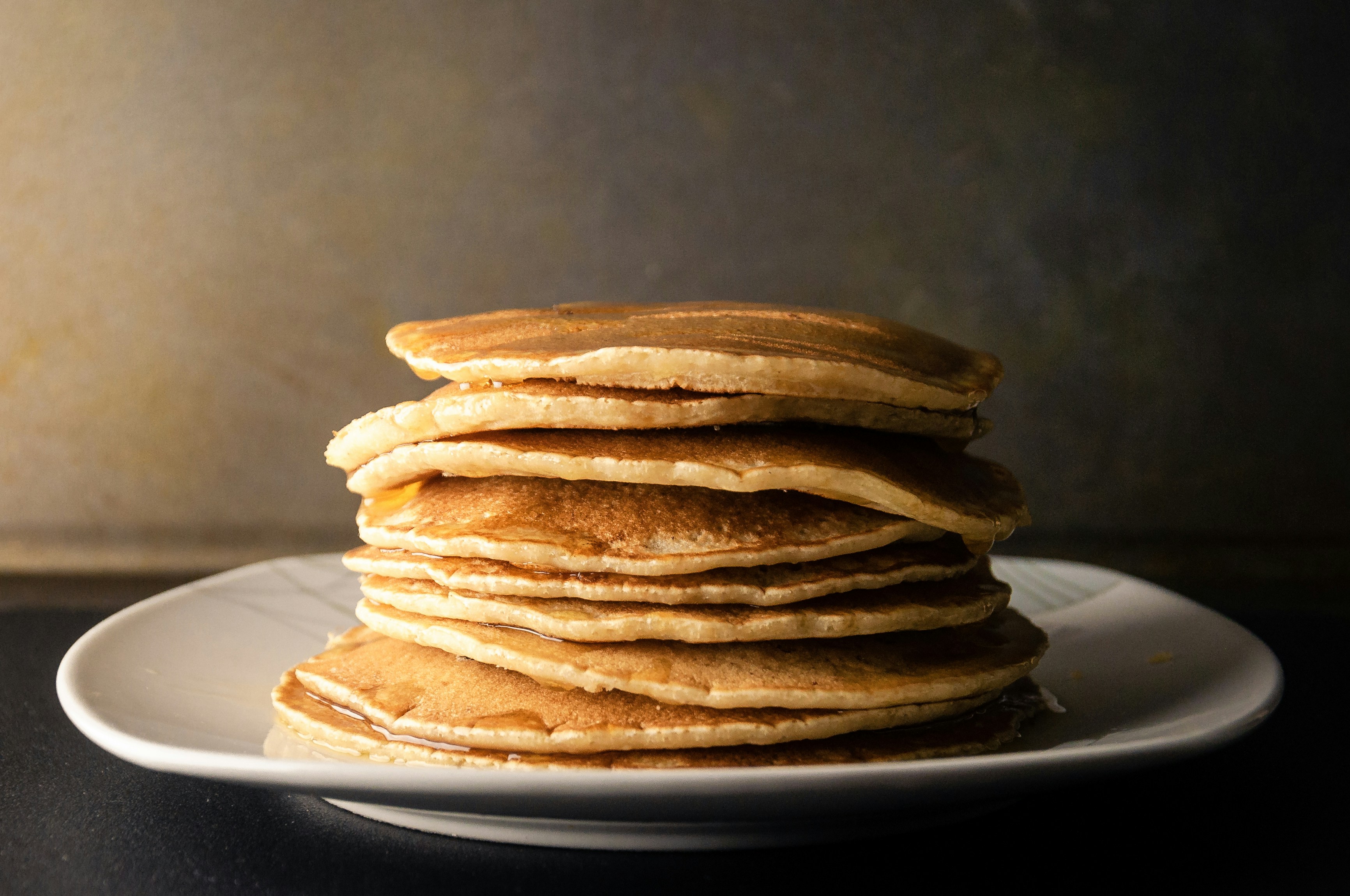 A neatly arranged stack of golden pancakes on a white plate, highlighting their fluffy texture against a softly lit background.