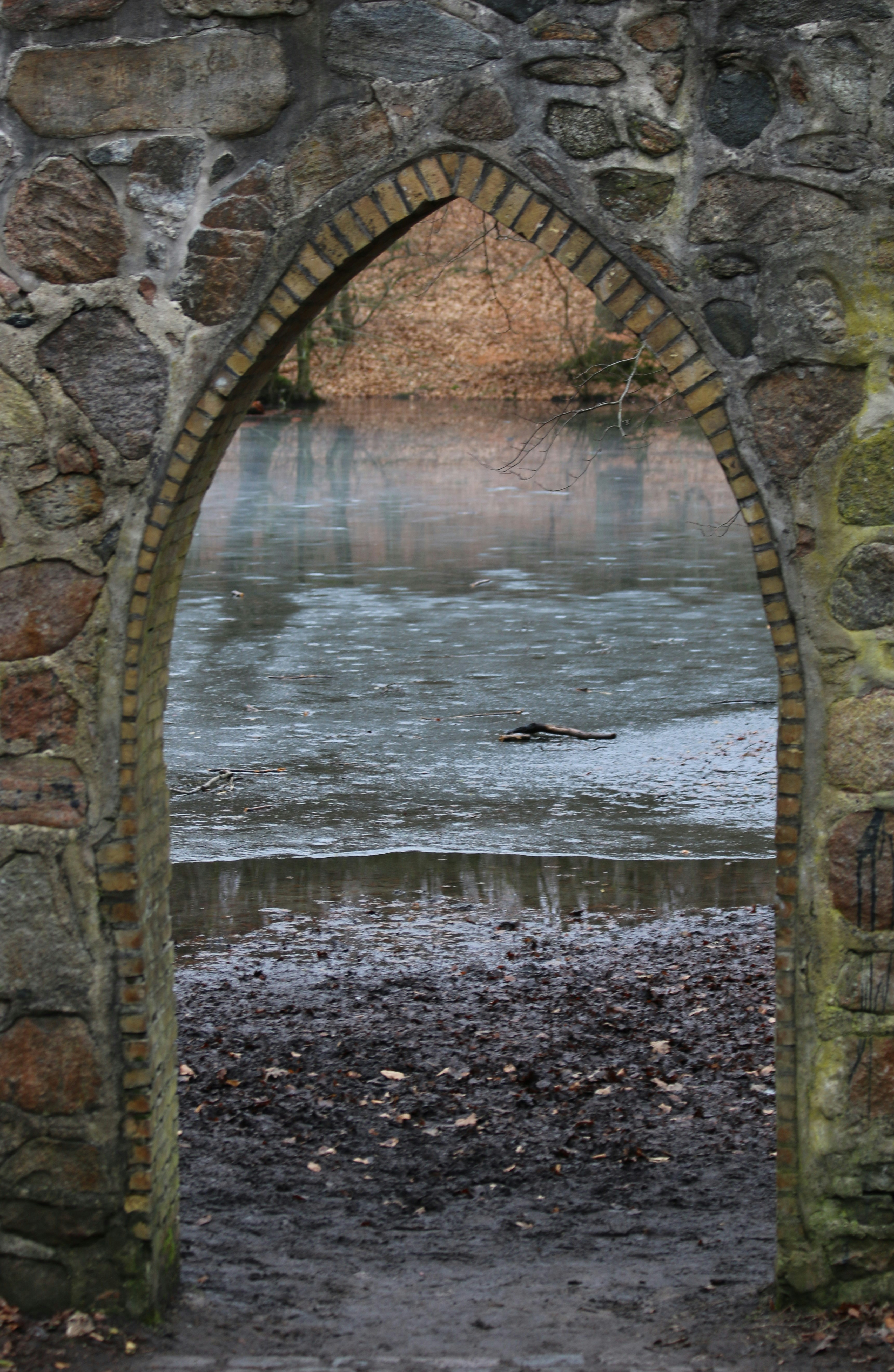 Stone archway leading to a serene, partially frozen pond surrounded by autumn foliage.