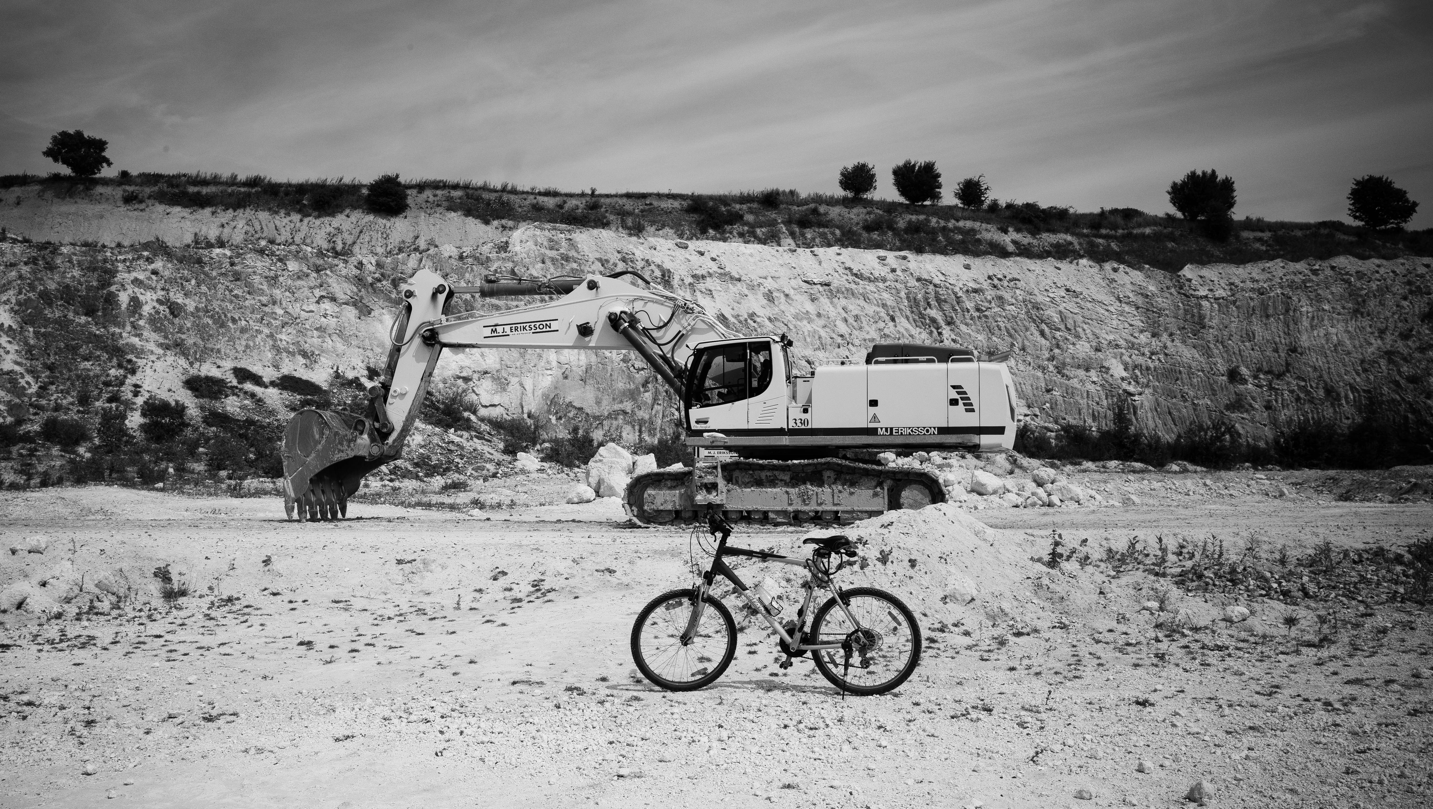 A bicycle stands in the foreground of an industrial landscape, dominated by a large excavator in black and white. The stark contrast highlights the juxtaposition of human leisure against heavy machinery.
