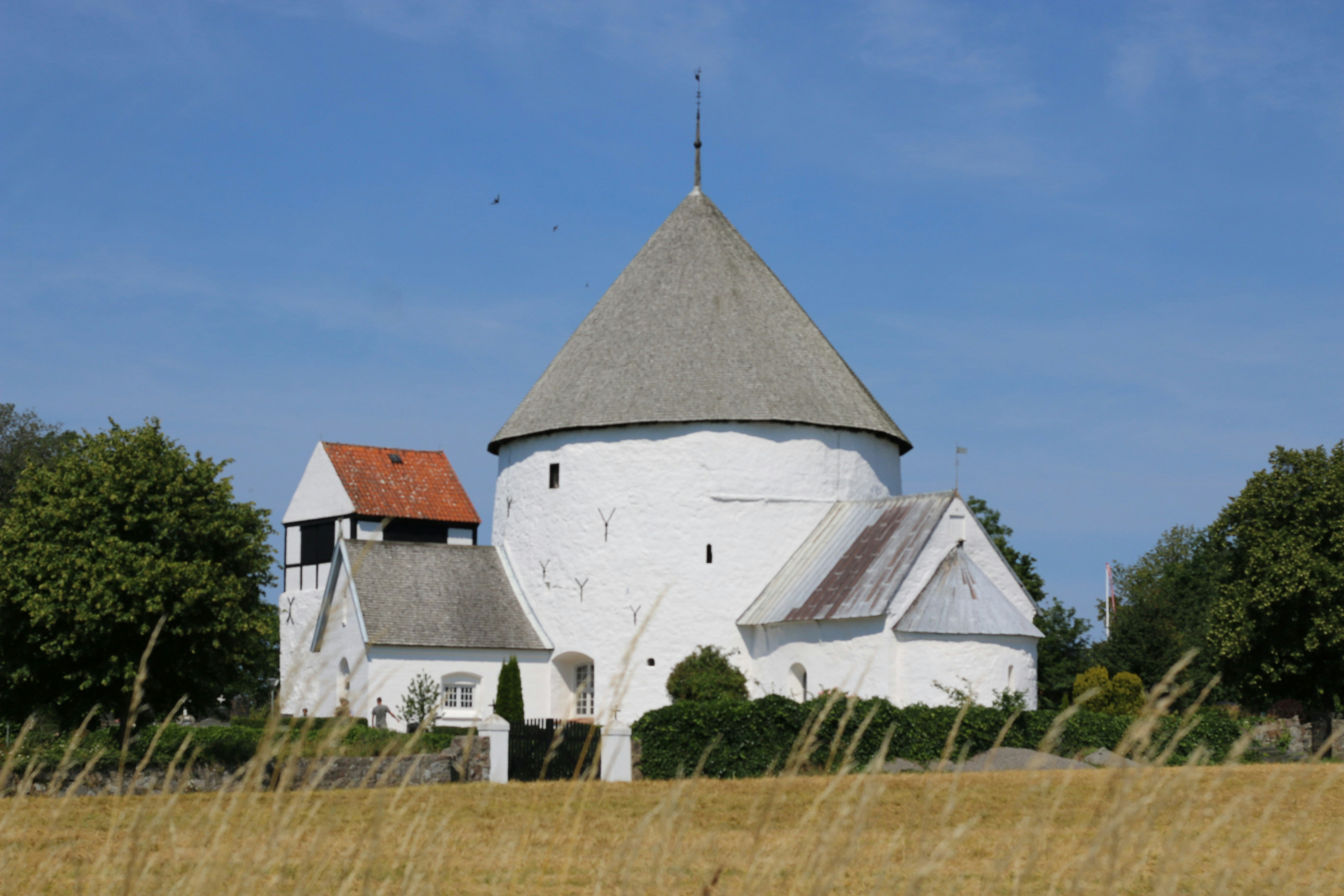 Historic round church with a conical roof, surrounded by lush greenery and golden fields under a clear blue sky.