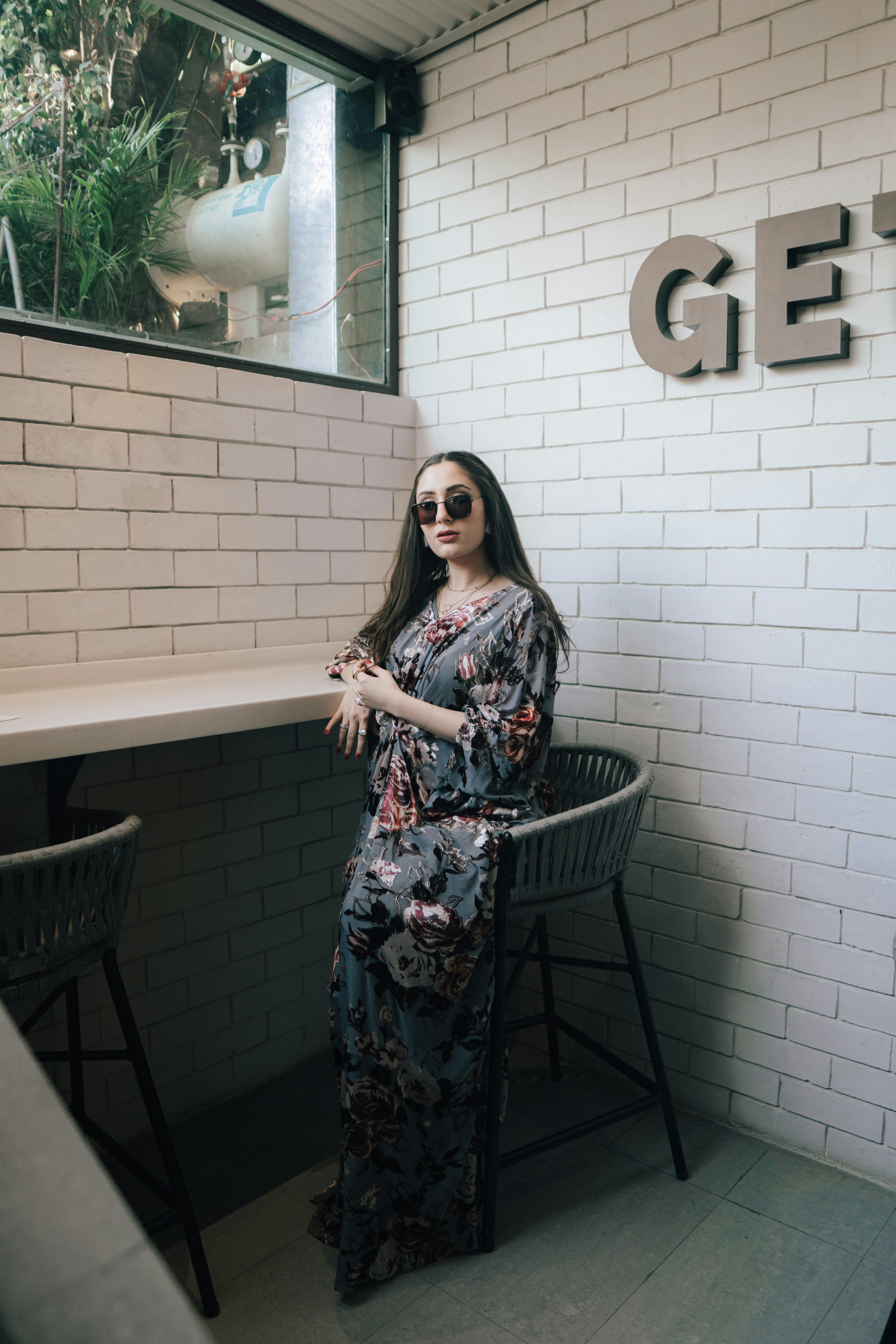 Woman in floral dress and sunglasses standing by a white brick wall with modern decor.