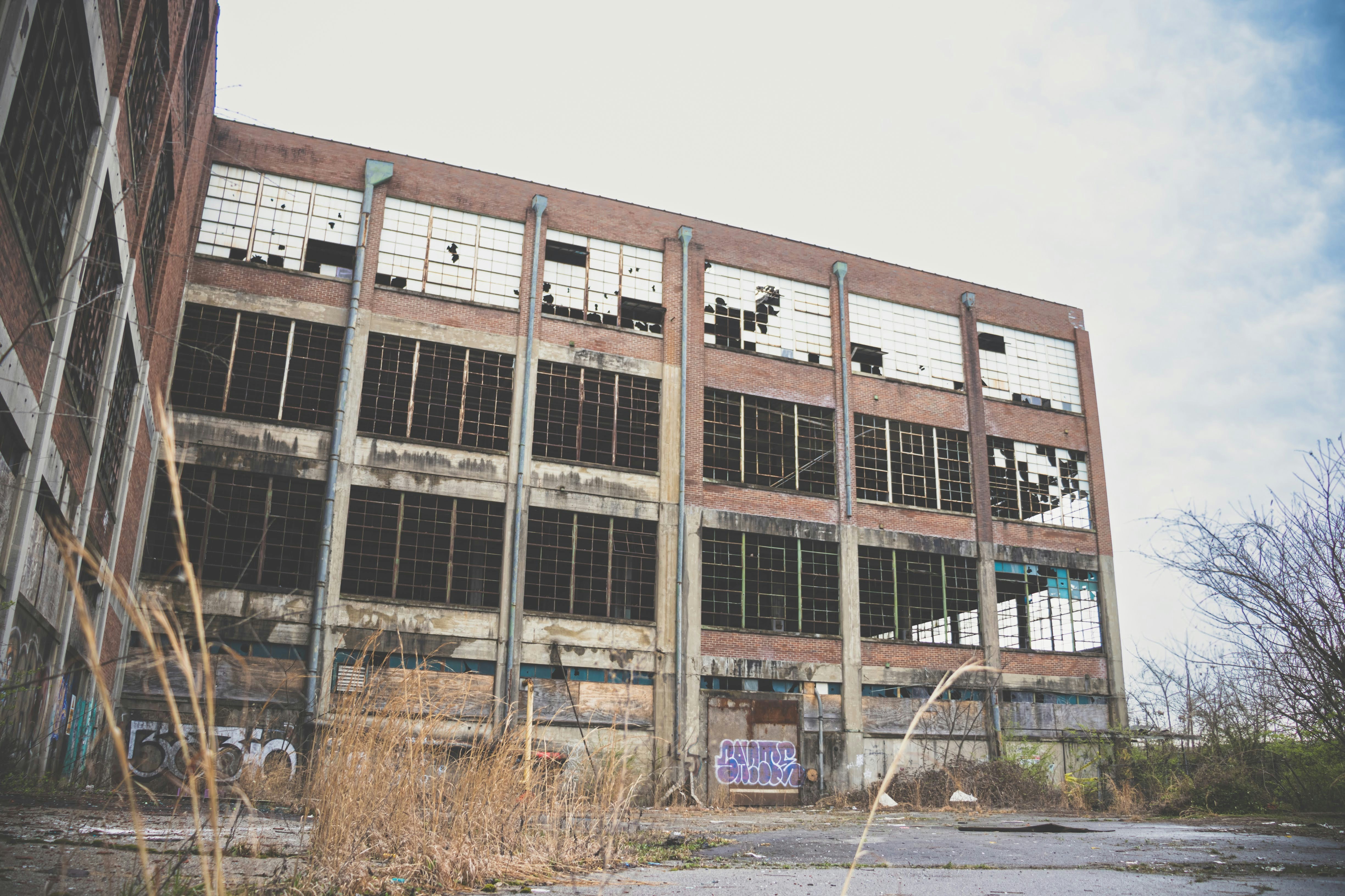 Abandoned industrial building with broken windows and graffiti, surrounded by overgrown grass and weeds.