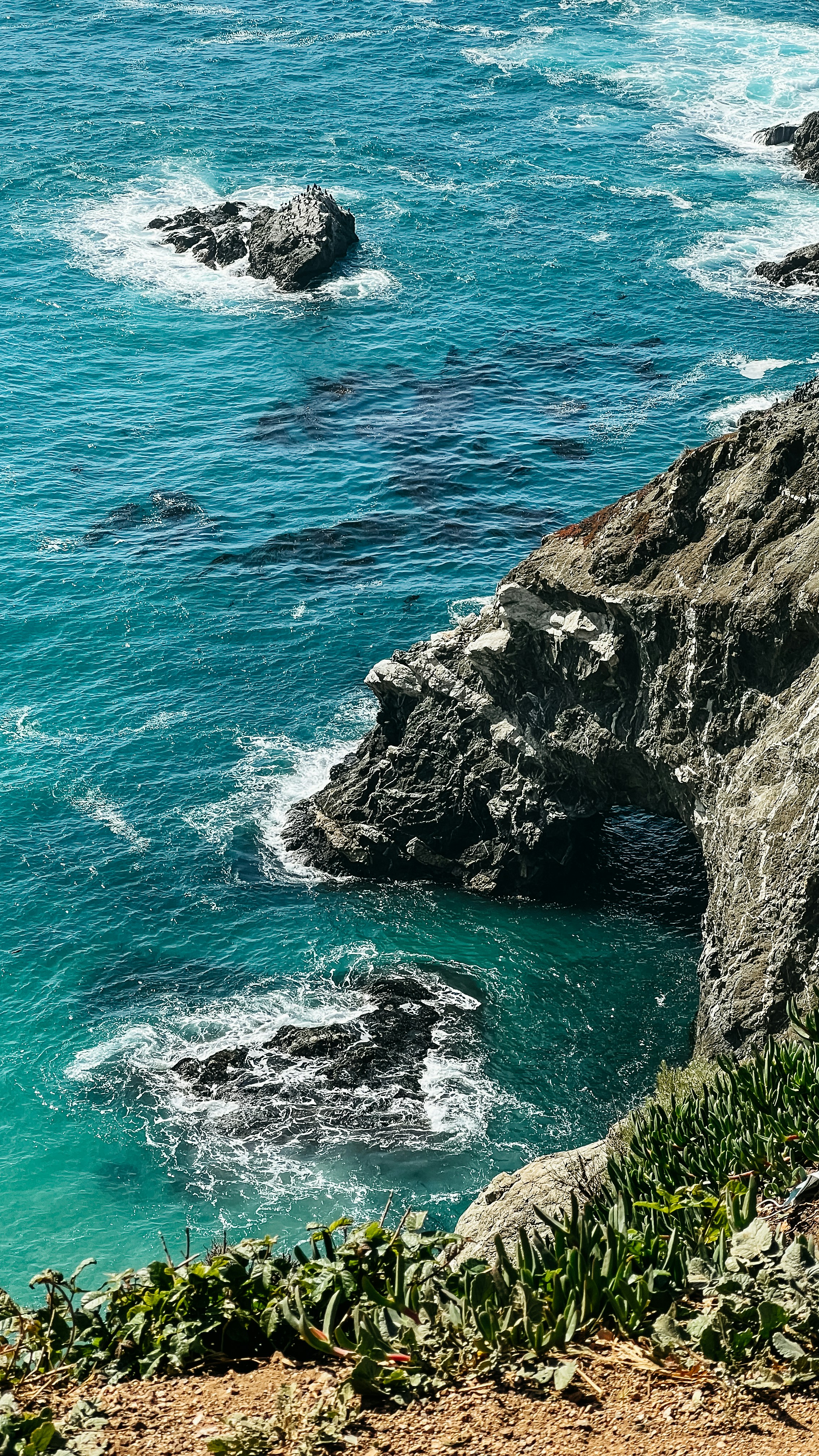 Rugged cliffs meet vibrant turquoise waters, with waves crashing against rocky formations. The scene captures the raw beauty of the coastline.