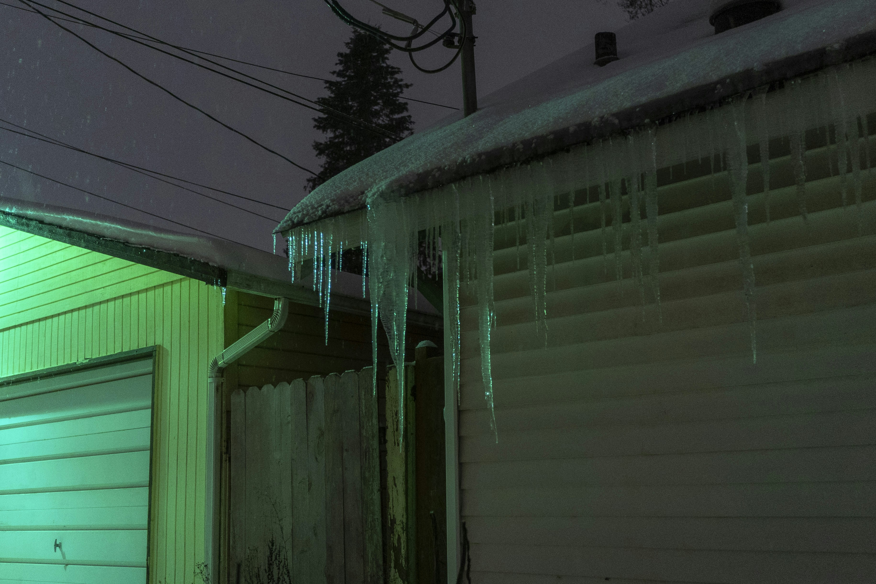Icicles hang from the eaves of a building, illuminated by a soft green light in a snowy alleyway at night.