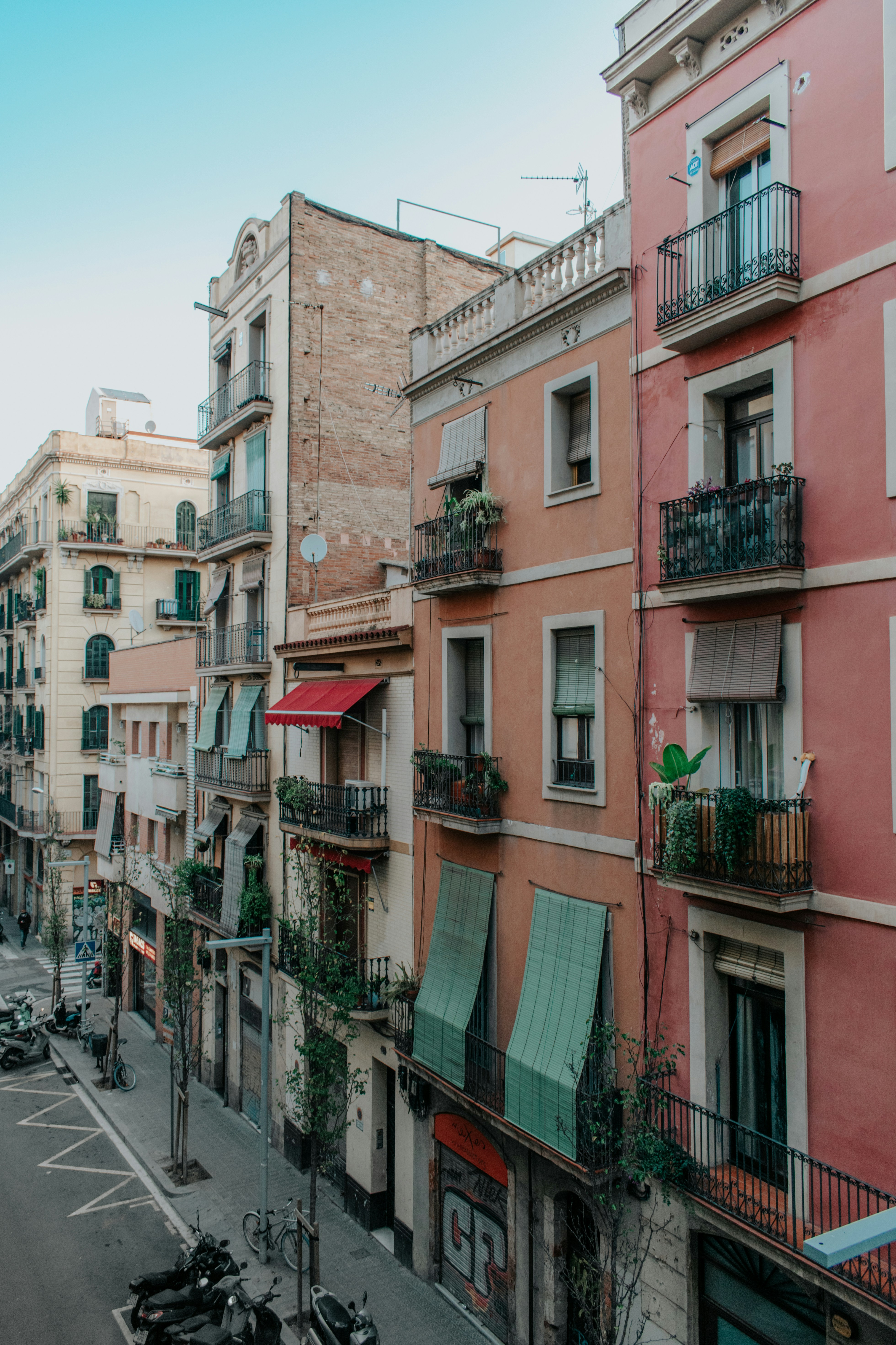 Colorful buildings line a quiet street in Barcelona, showcasing unique balconies and greenery. The scene captures the essence of urban life in a historic district.