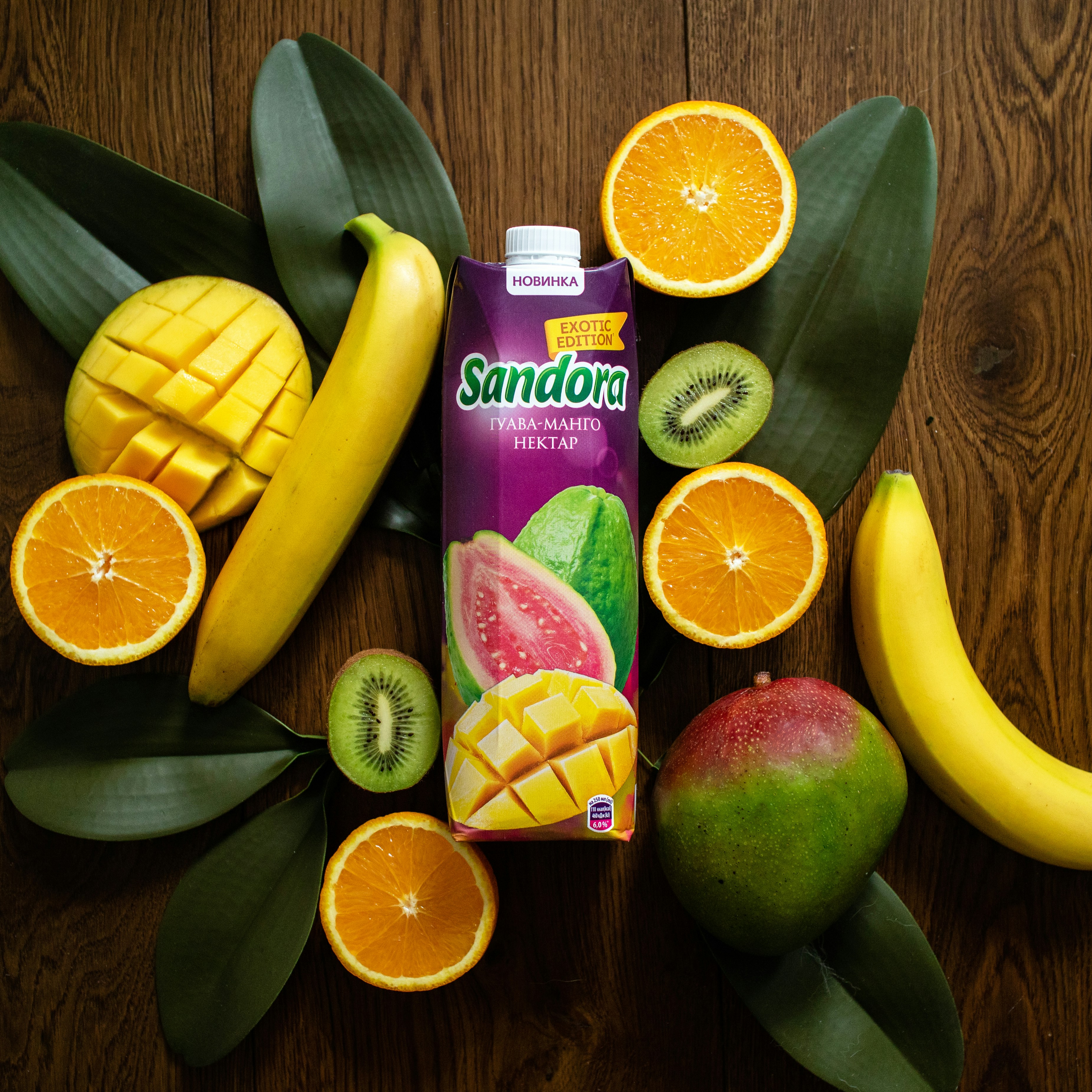 Product photograph of a Sandora mango nectar carton surrounded by sliced oranges, kiwis, a mango, and bananas on a wooden surface.