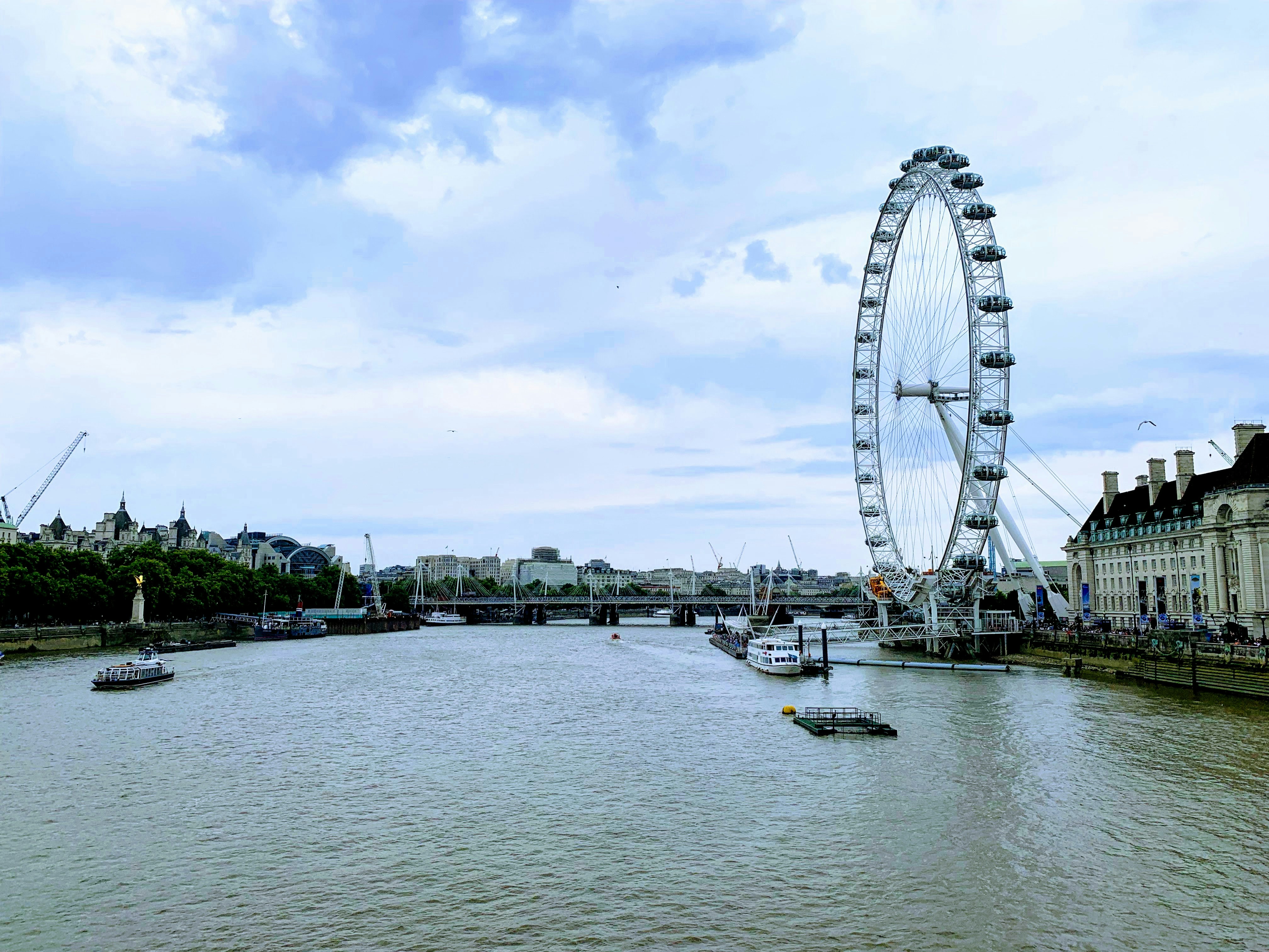 London Eye towering over the River Thames, framed by a mix of historic and modern architecture under a cloudy sky.