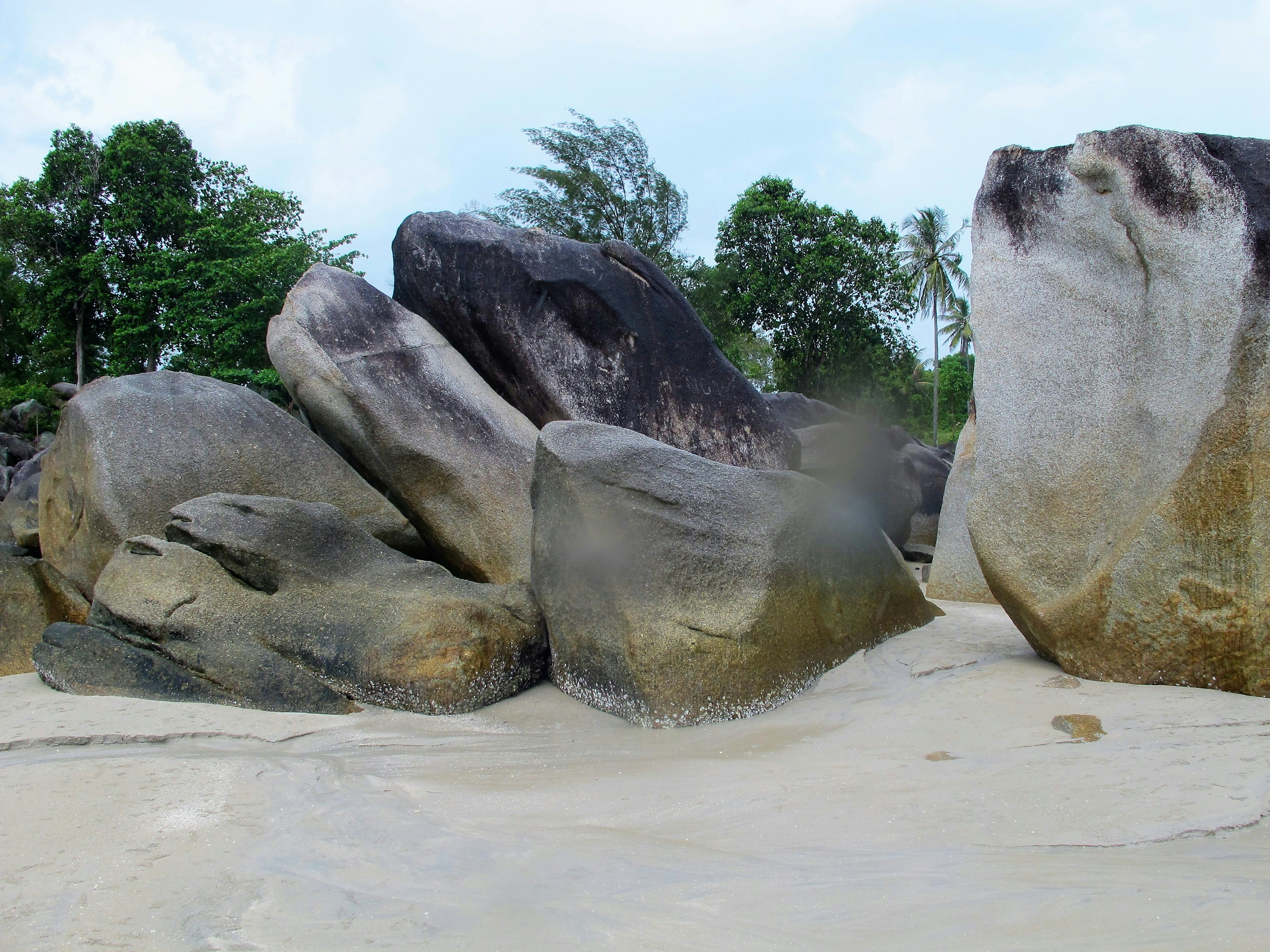 Massive boulders rise from sandy beach, surrounded by lush greenery under a cloudy sky.