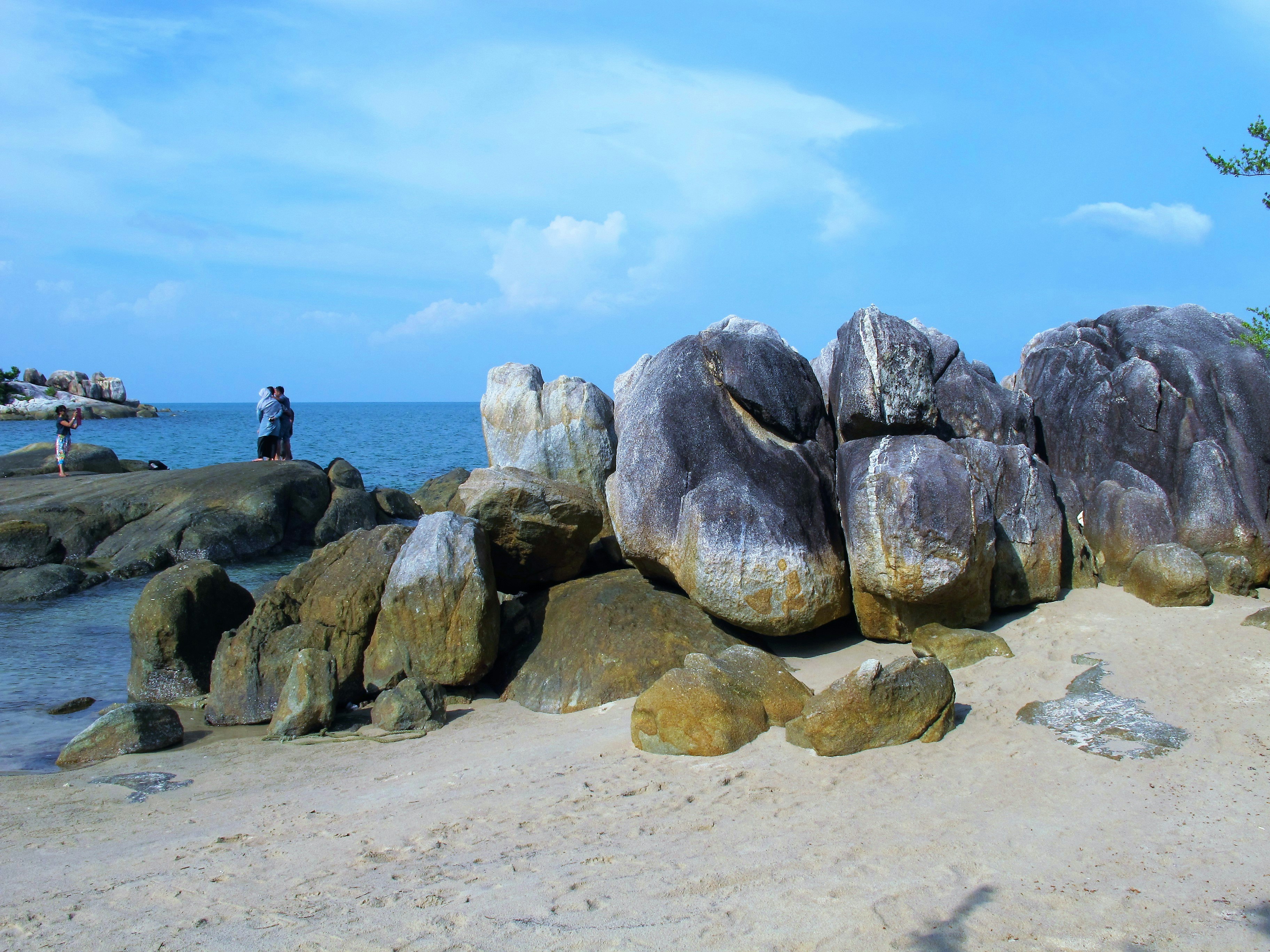 Large, textured boulders frame a tranquil beach scene with two figures exploring the shoreline. The serene ocean stretches into the distance.