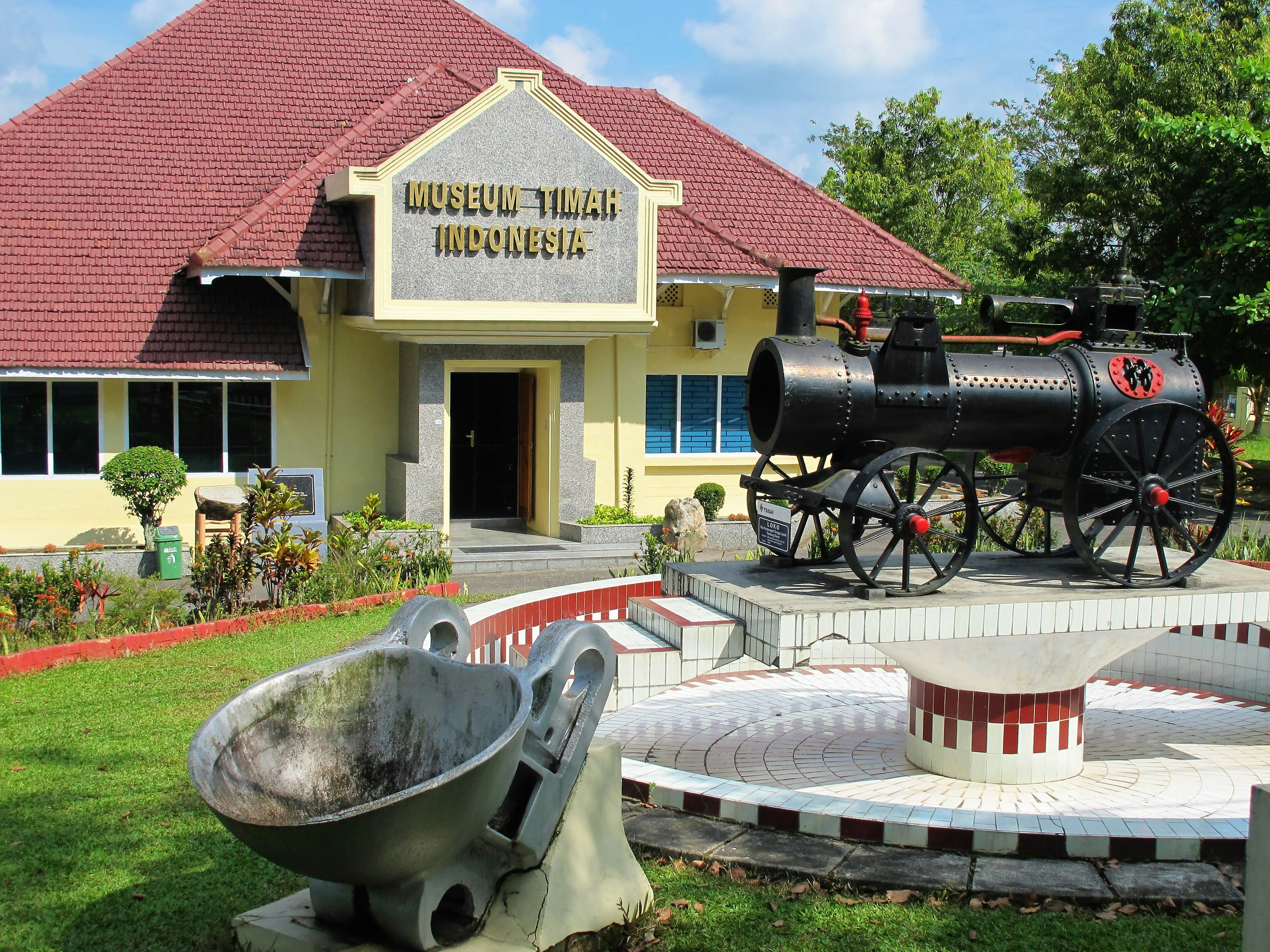 A vintage steam locomotive displayed prominently outside the Museum Timah Indonesia, surrounded by lush greenery and historical artifacts.