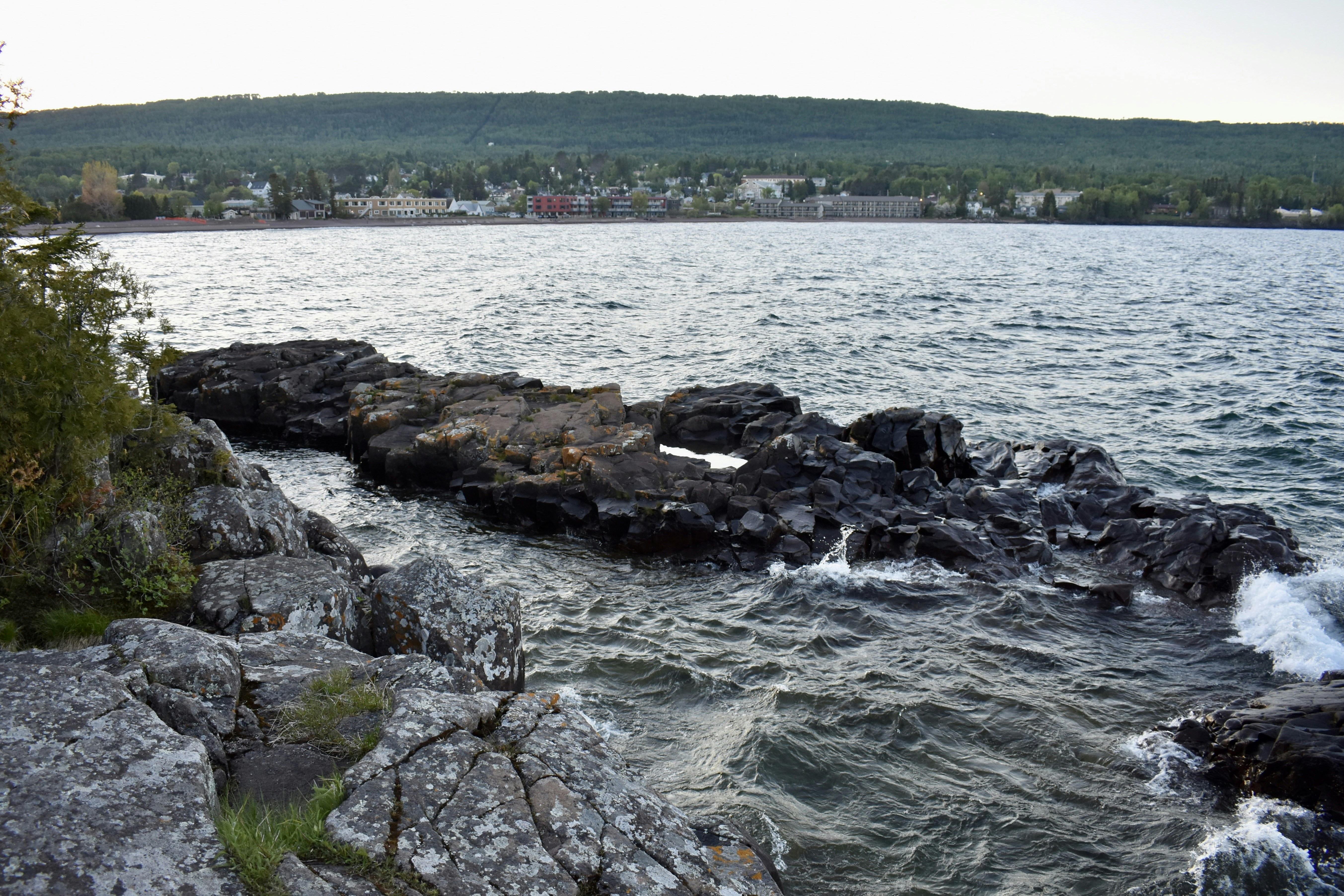 Rugged rocks jutting into a calm lake, framed by lush greenery and a distant town under a soft sky.