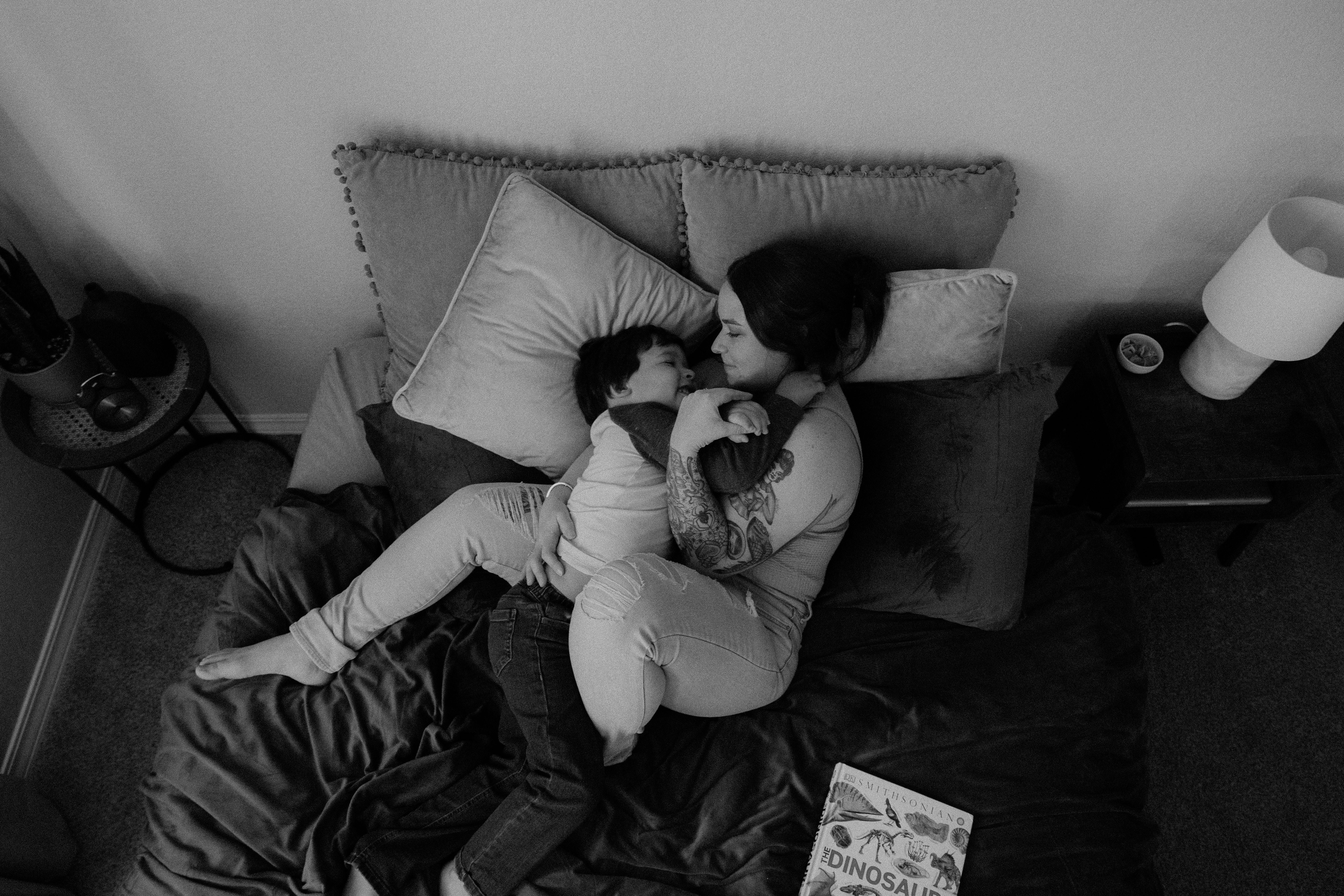 A mother and child share a loving embrace on a plush bed, surrounded by soft pillows and blankets. A book lies nearby, suggesting a moment of shared storytelling.