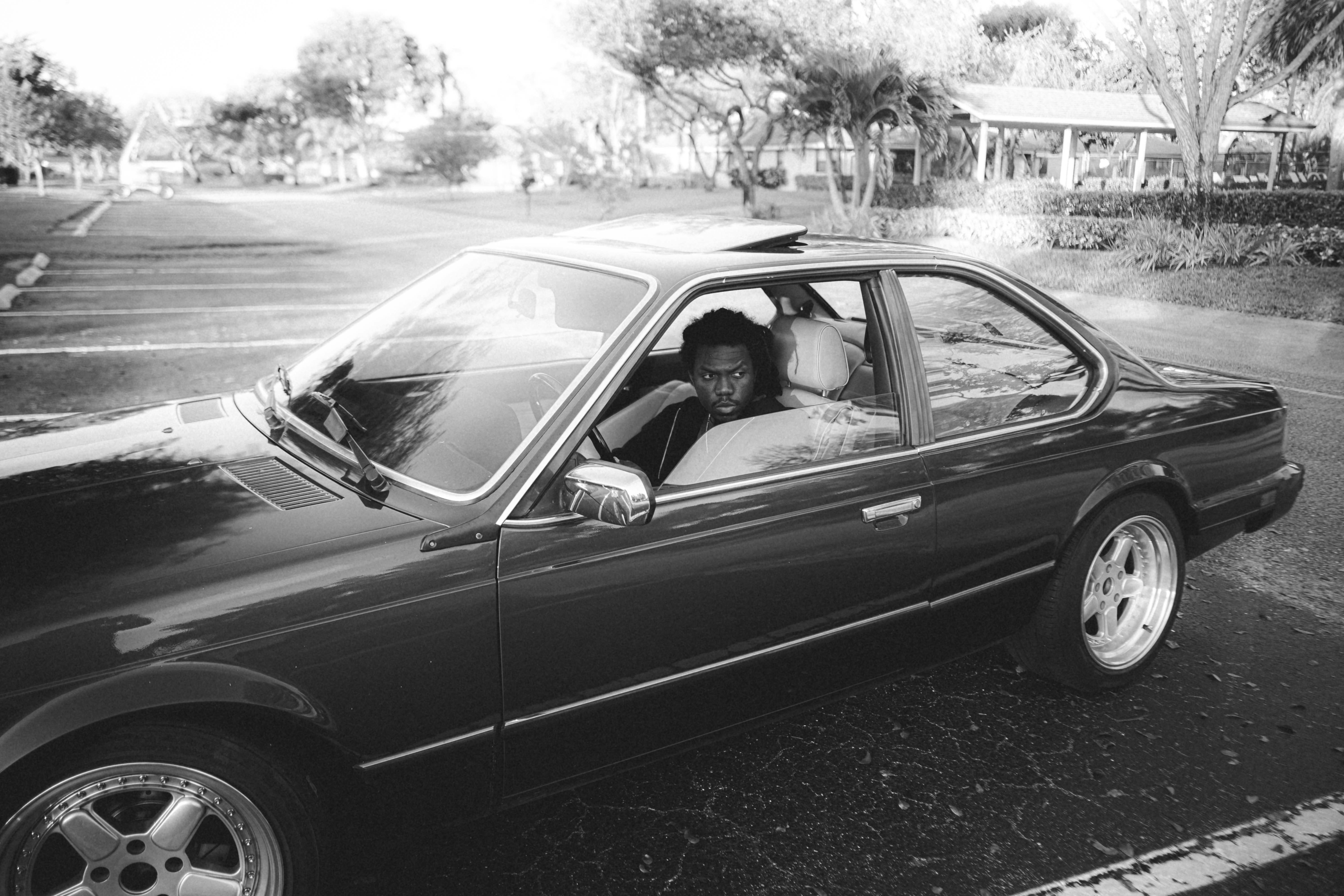 A driver gazes thoughtfully from the interior of a classic car parked in an empty lot, surrounded by trees. The monochromatic tones enhance the mood of contemplation.