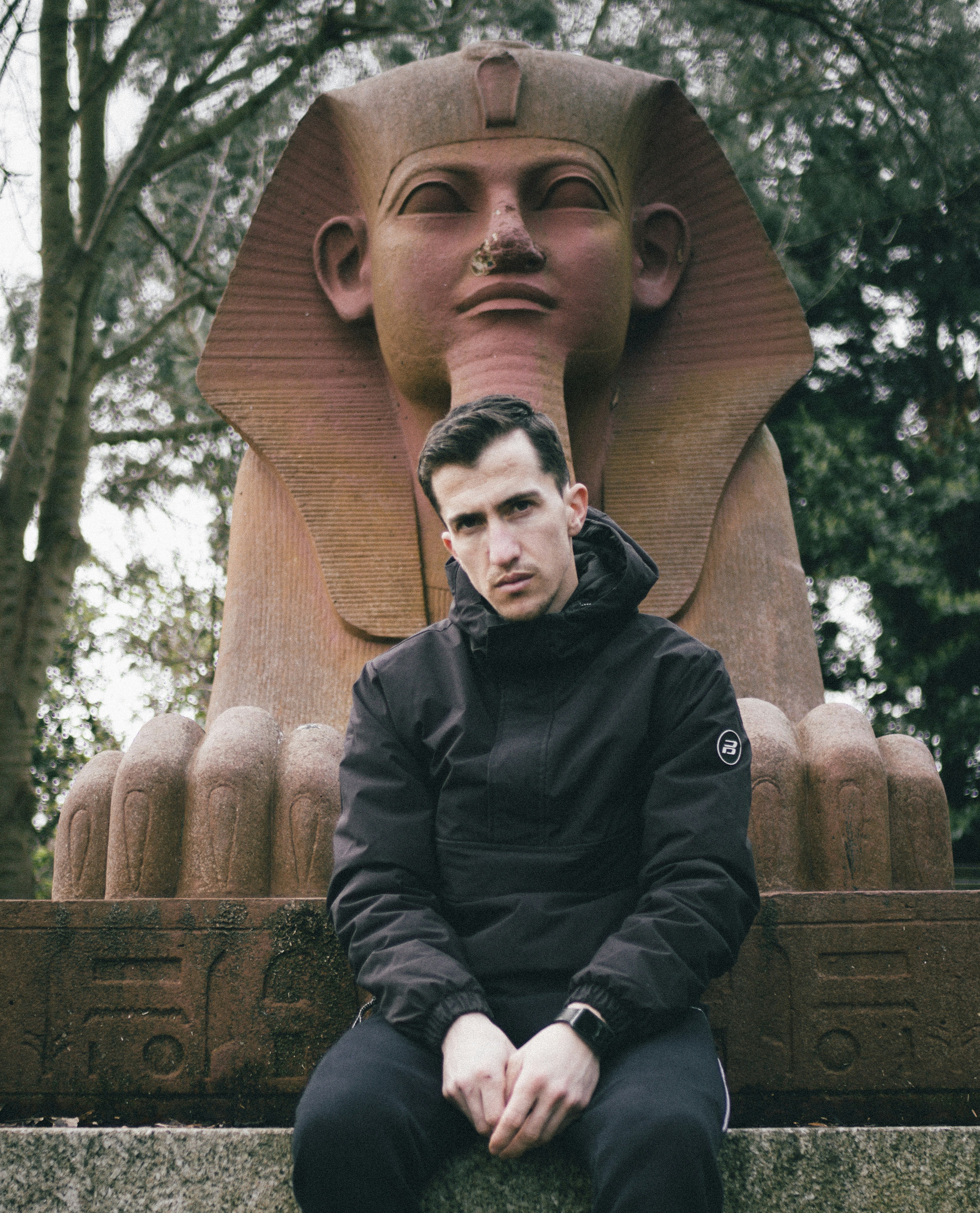 A young man seated in front of a monumental sphinx sculpture, exuding a blend of ancient history and contemporary style.