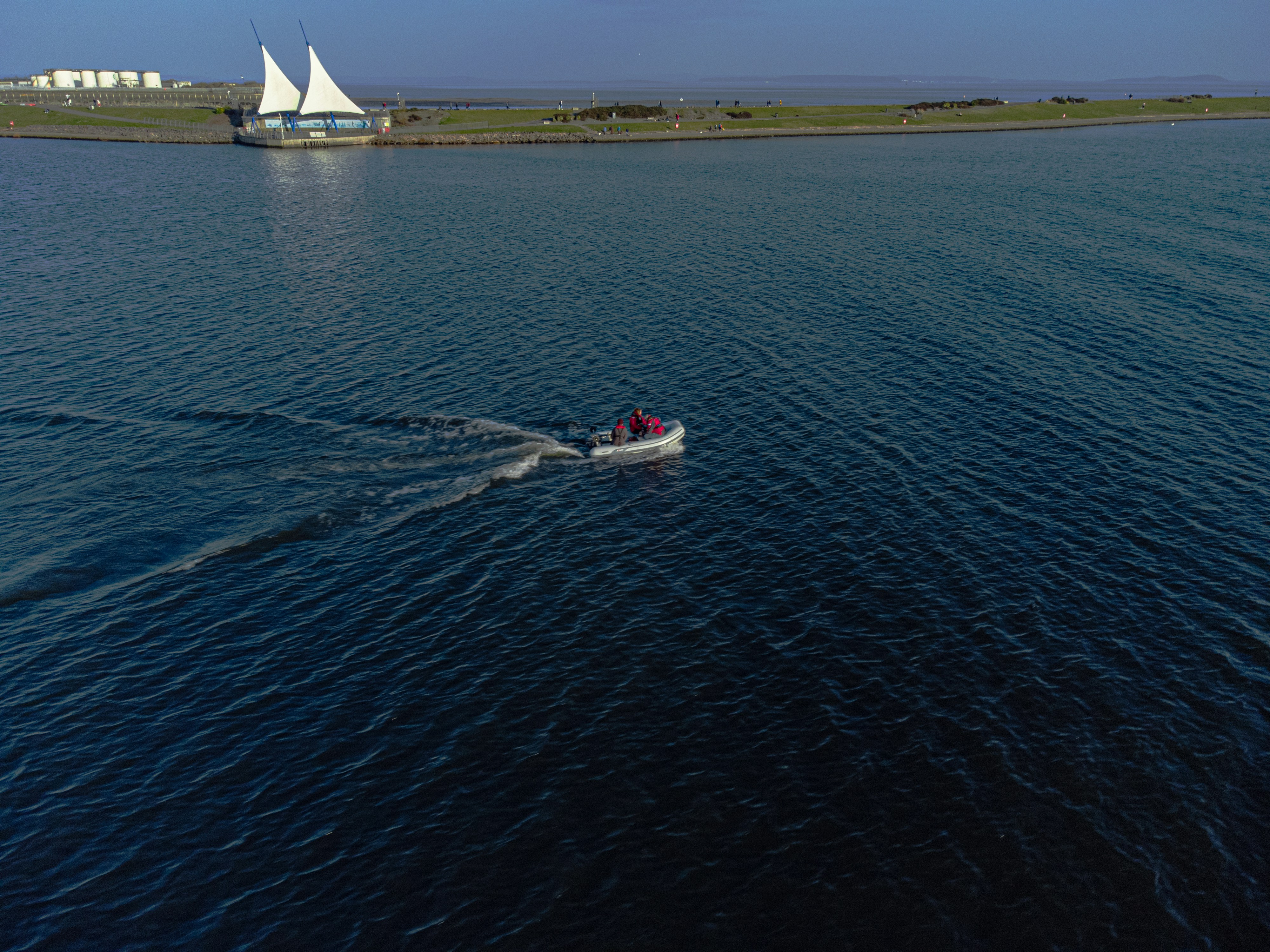 Drone photograph of a small motorboat carving a wake through deep blue water toward a distant shoreline where a white sailboat rests near low buildings.