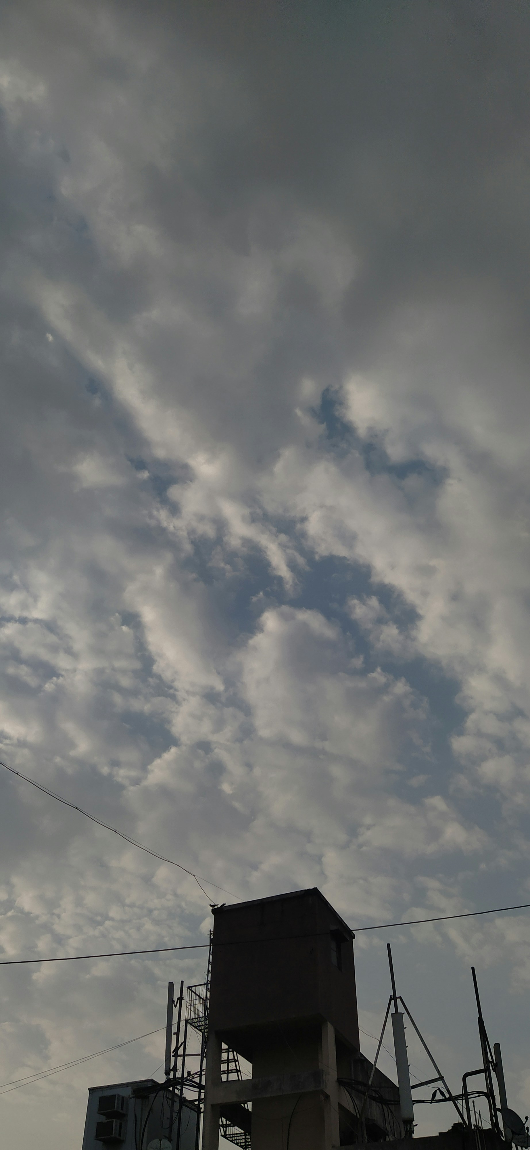 Dramatic cloud formations hover above a cityscape with a water tank and communication structures. The interplay of light and shadow creates a moody atmosphere.