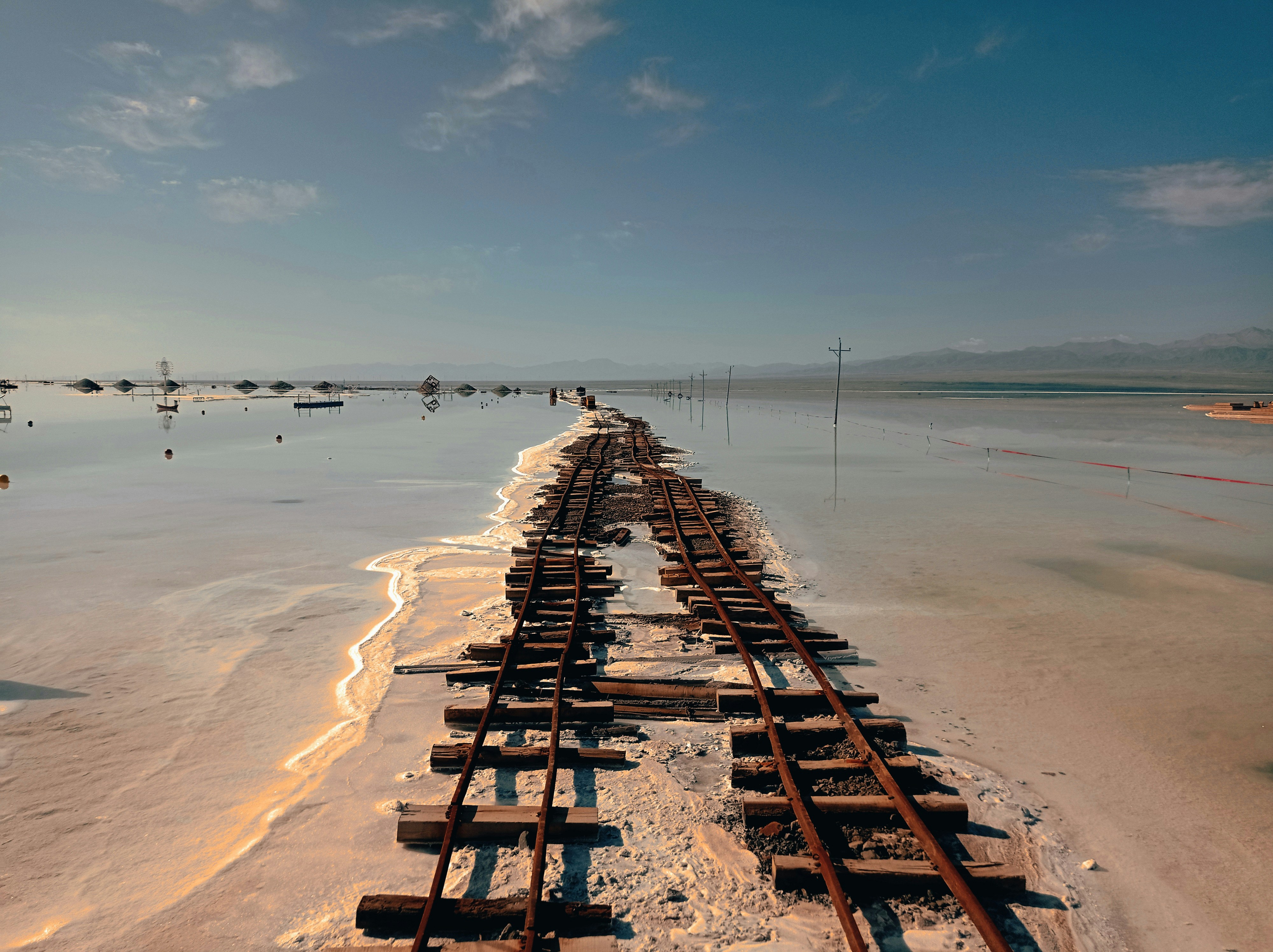 Abandoned railway tracks extend into a serene, shallow body of water, reflecting the clear sky above. The juxtaposition of rusted metal and tranquil water creates a mesmerizing scene.
