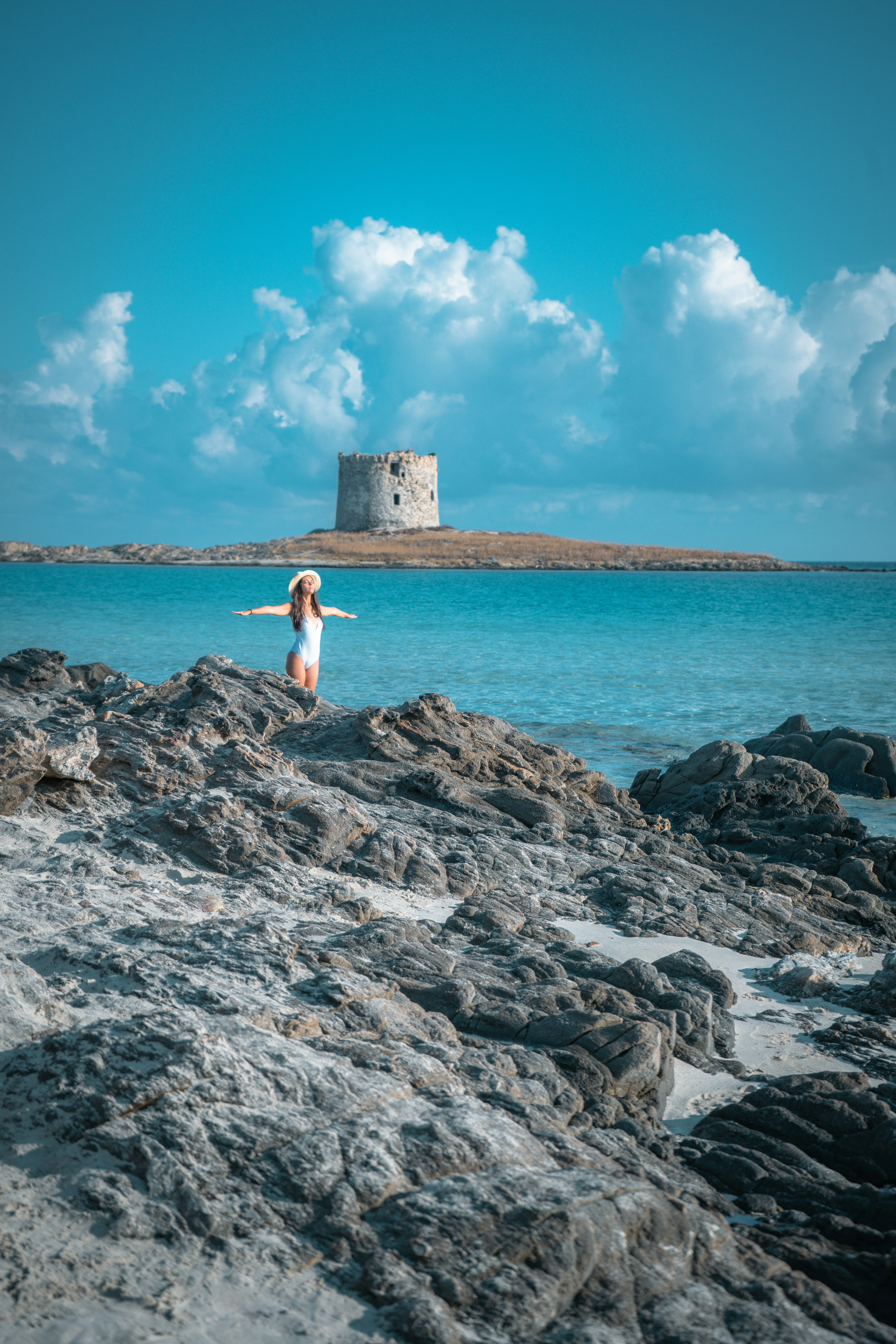 Turquoise Promenade: A Lone Figure, Rocky Foreground, and a Distant Island Tower