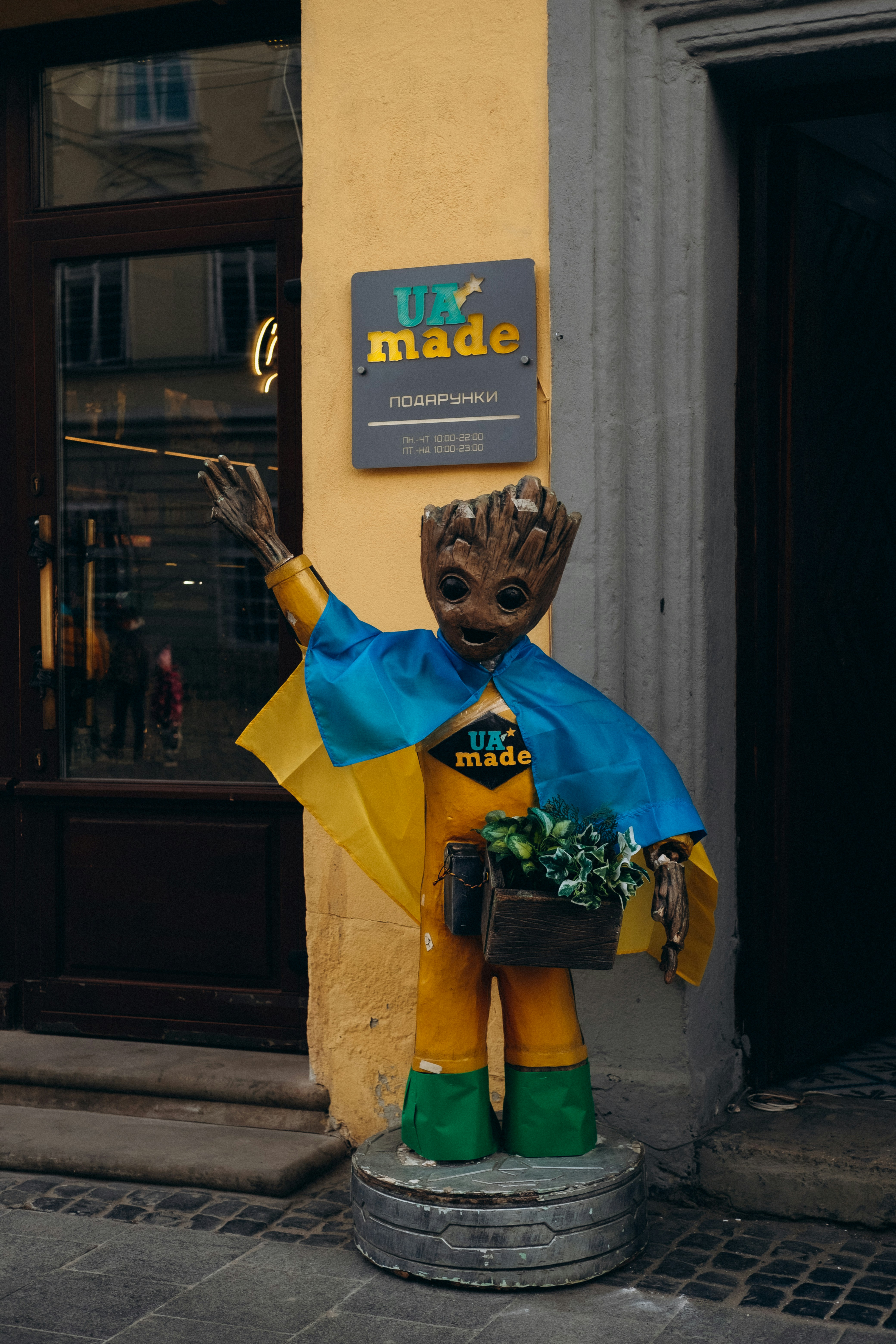 Whimsical wooden figure dressed in blue and yellow, holding a basket of plants, stands outside a shop with a sign promoting local products.