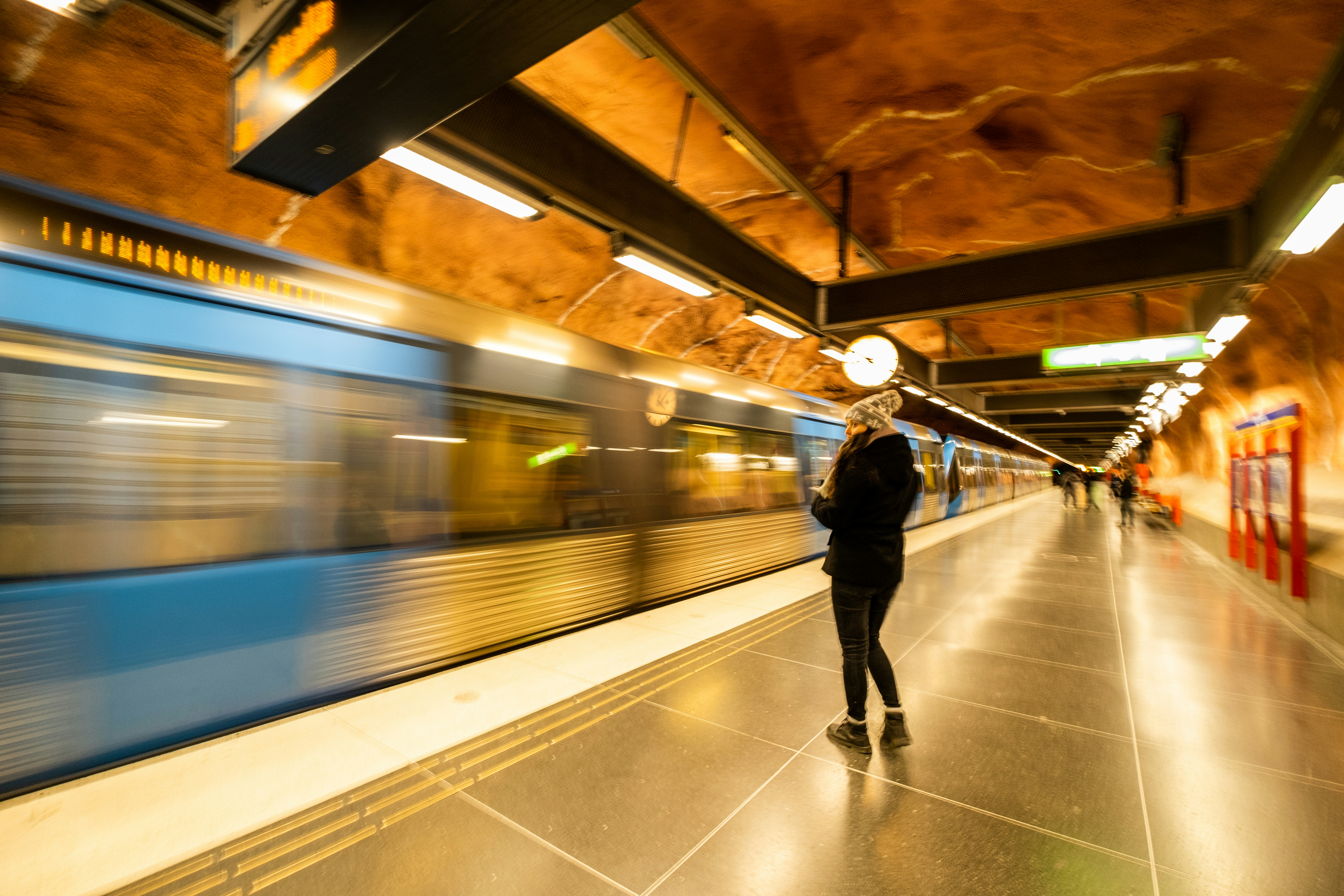 A person stands still on a subway platform as a blue train rushes past, creating a dynamic contrast between motion and stillness.