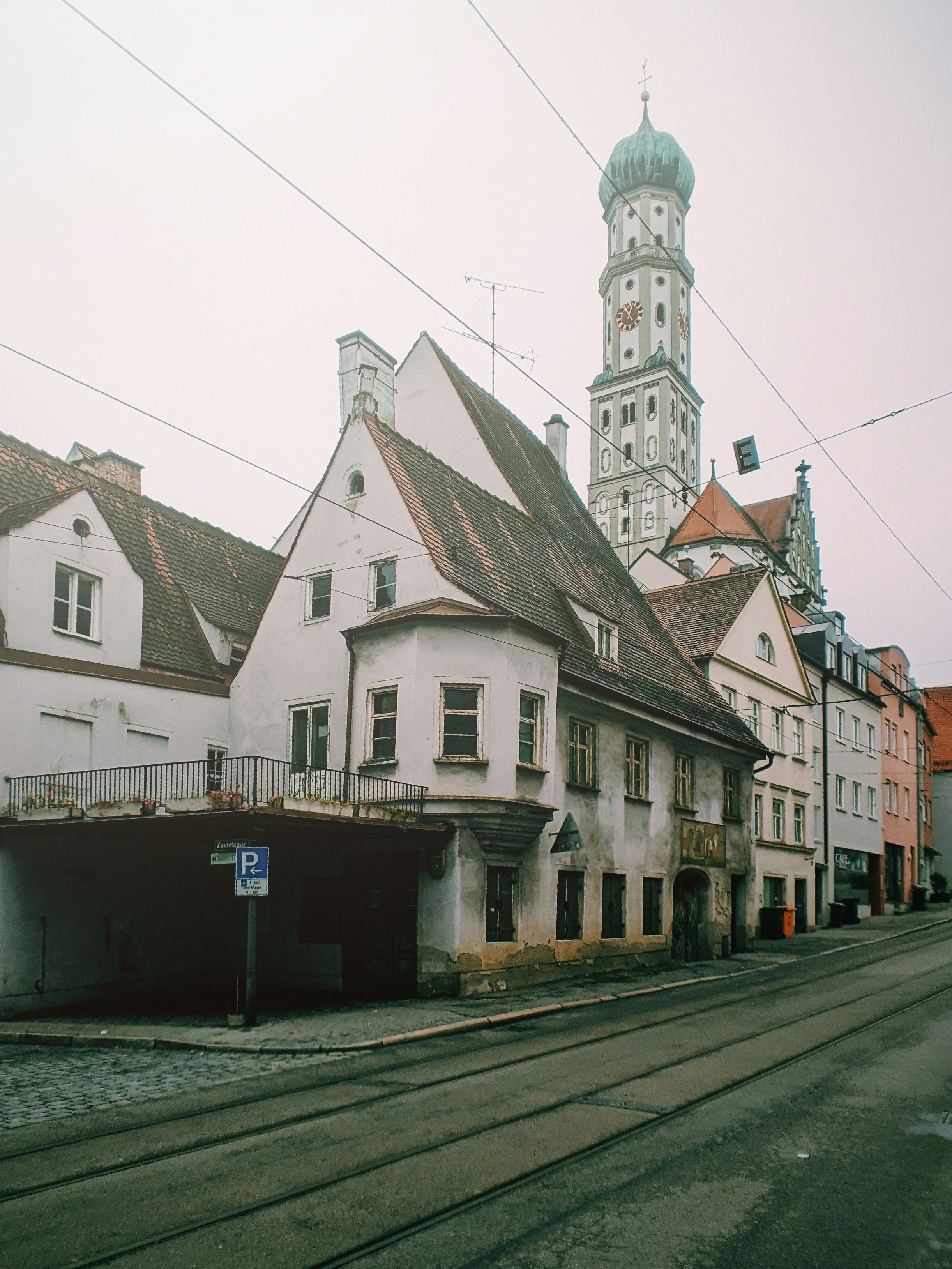 Historic buildings lining a cobblestone street in Augsburg, with a prominent clock tower in the background. The scene captures the charm of old-world architecture.