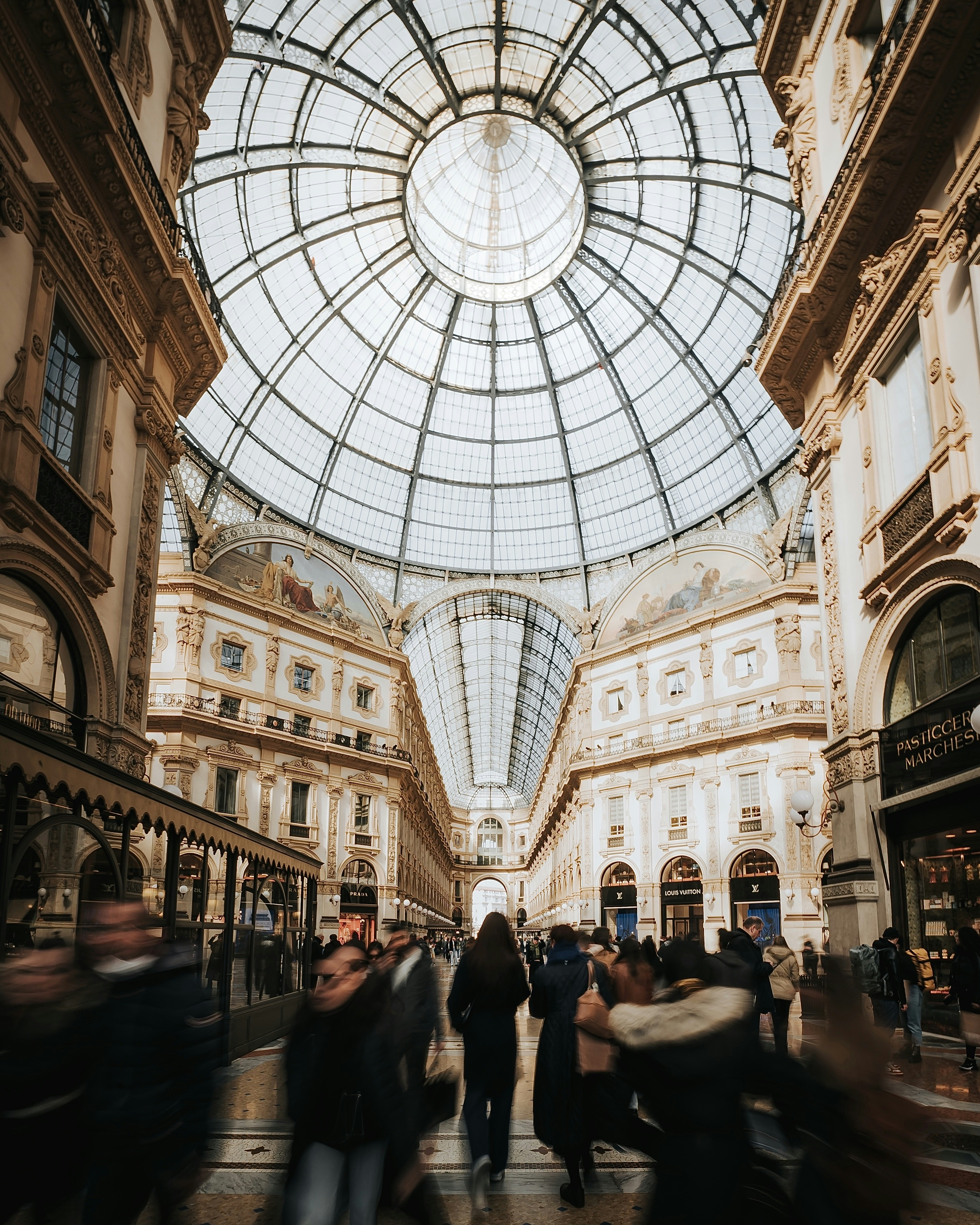 Interior of a historic gallery showcasing intricate architecture and a stunning glass dome, filled with bustling visitors.
