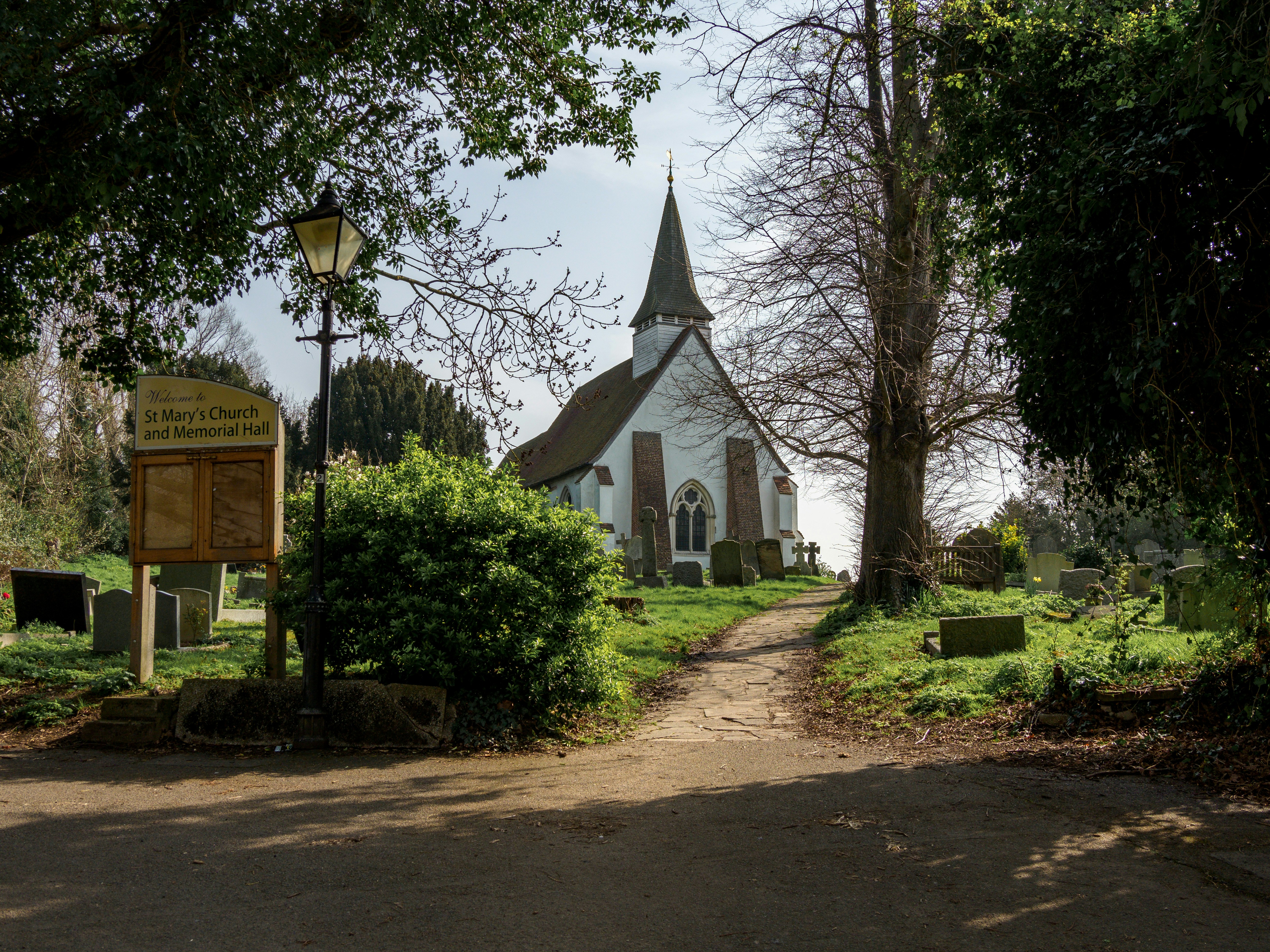 Charming church nestled among greenery, with a path leading to its entrance, surrounded by gravestones. A vintage lamp post adds to the tranquil ambiance.