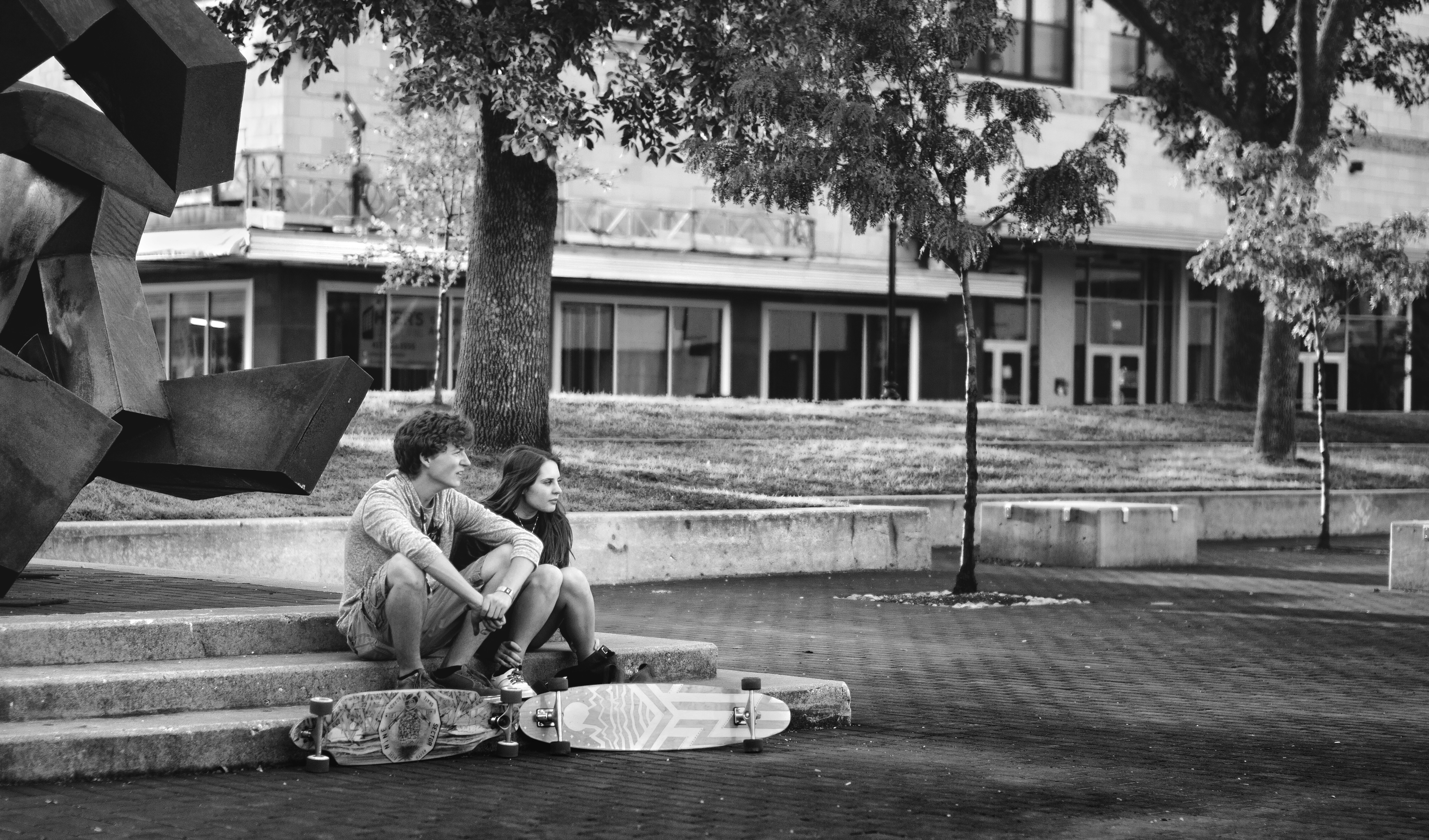Two young people sit on steps with skateboards, next to a modern sculpture in an urban setting.