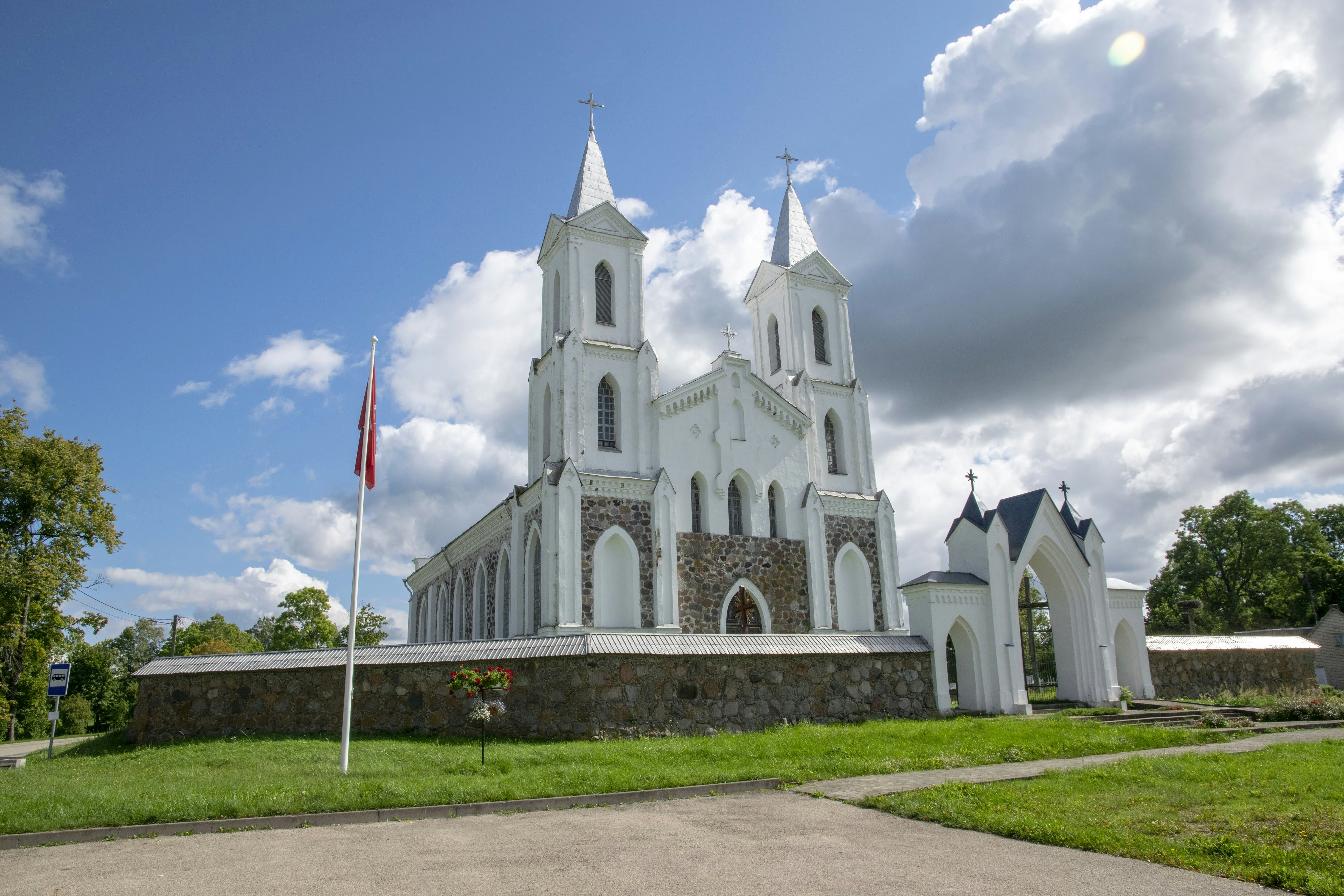 A striking white church with twin spires stands against a backdrop of fluffy clouds, showcasing intricate stonework and surrounding greenery.