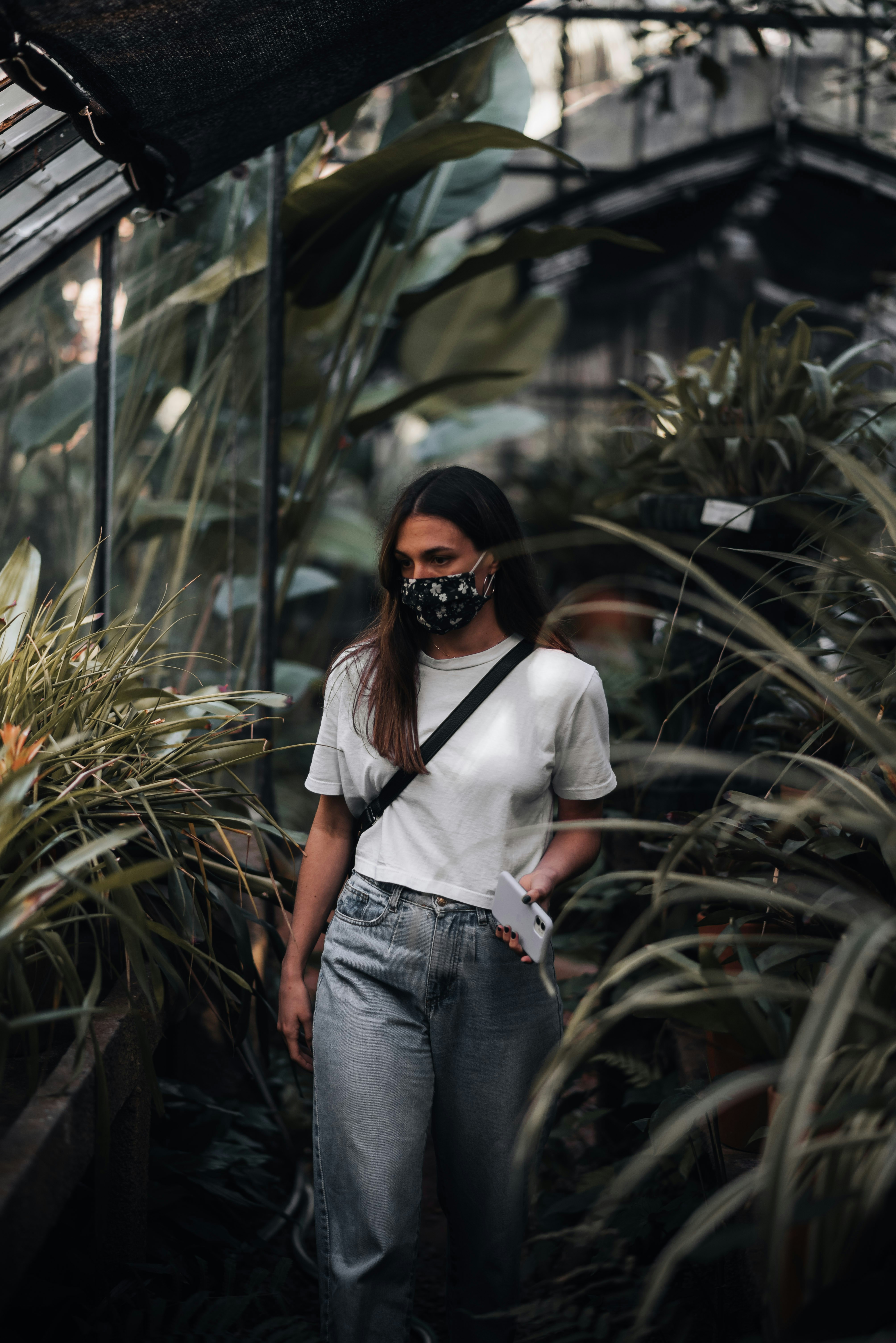 A woman in a patterned mask strolls through a lush greenhouse, surrounded by vibrant plants and foliage.