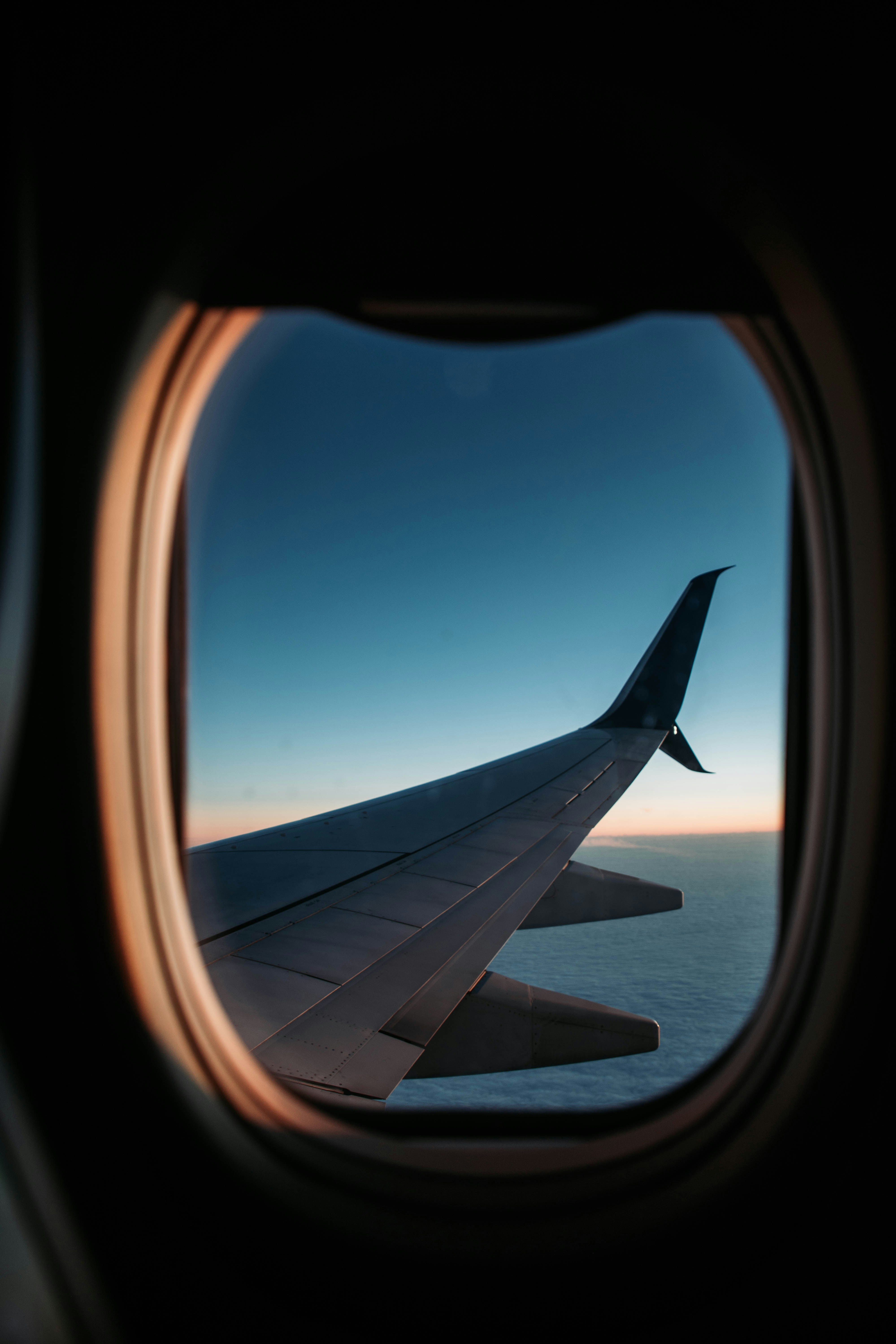 Airplane wing extending into the horizon, framed by an aircraft window, showcasing a serene sky transitioning from day to night.